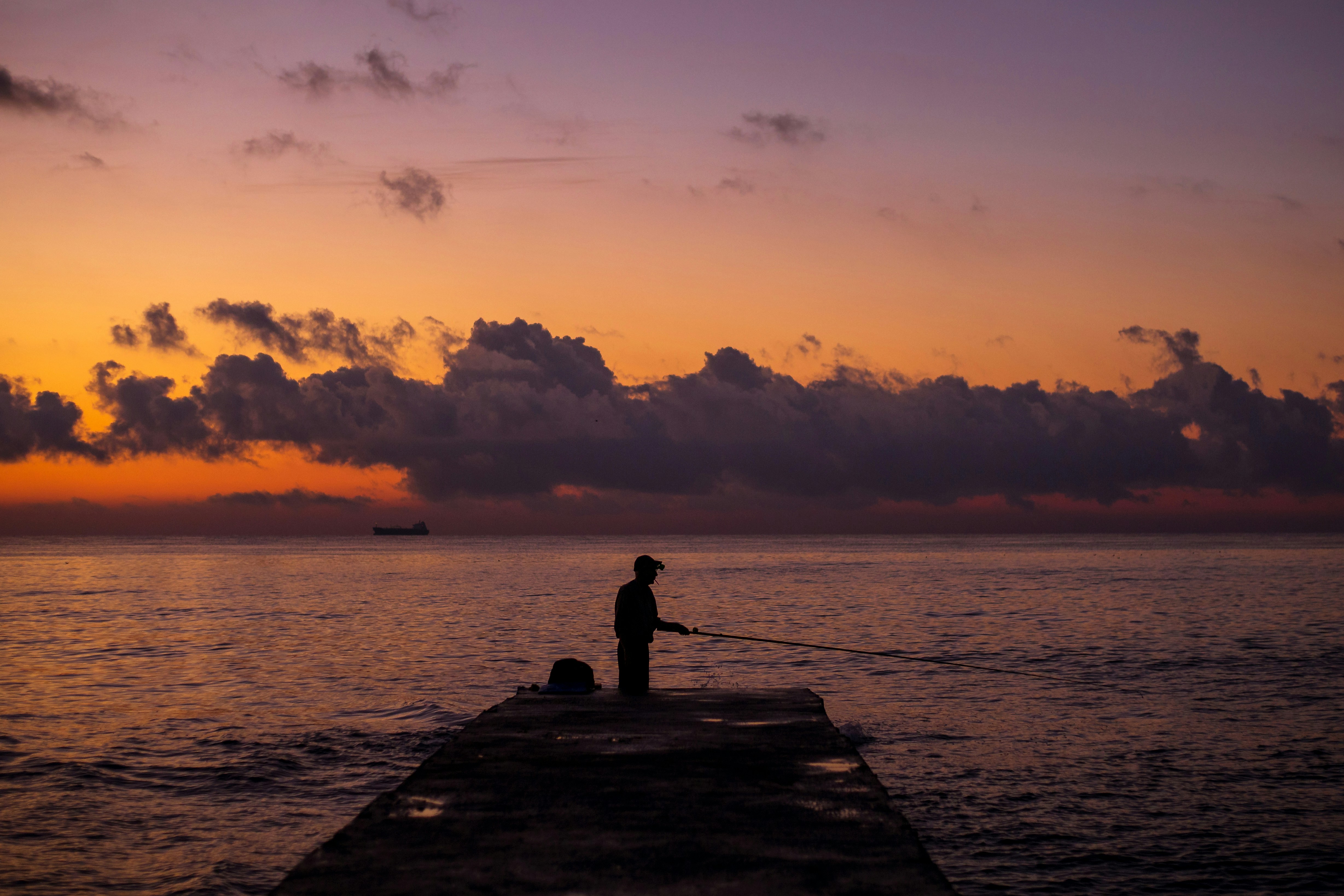 Silhouette of a fisherman on a pier against a vibrant sunrise sky over calm ocean waters.