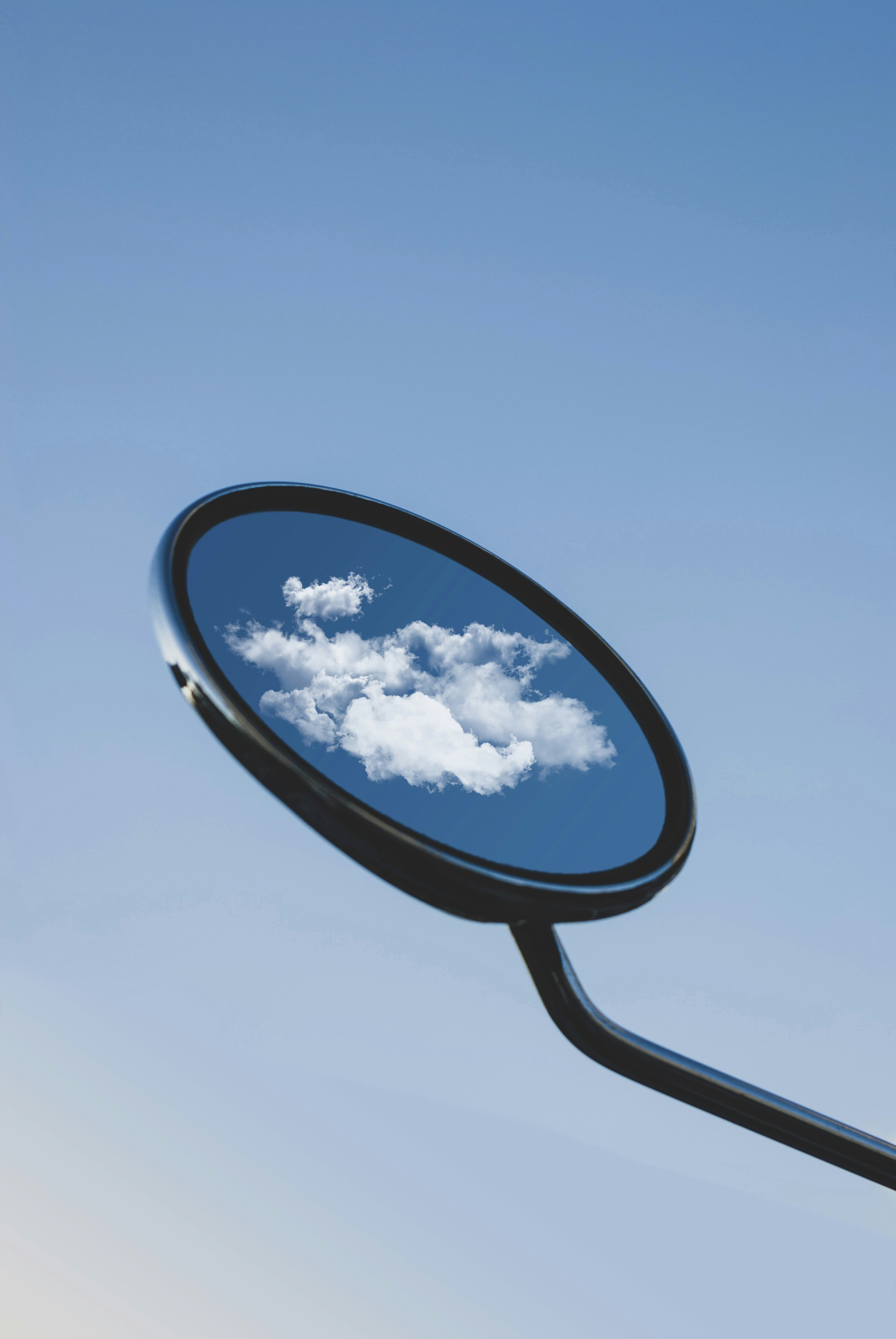 Circular mirror reflecting blue sky and clouds, mounted on a curved black arm against a clear gradient backdrop.