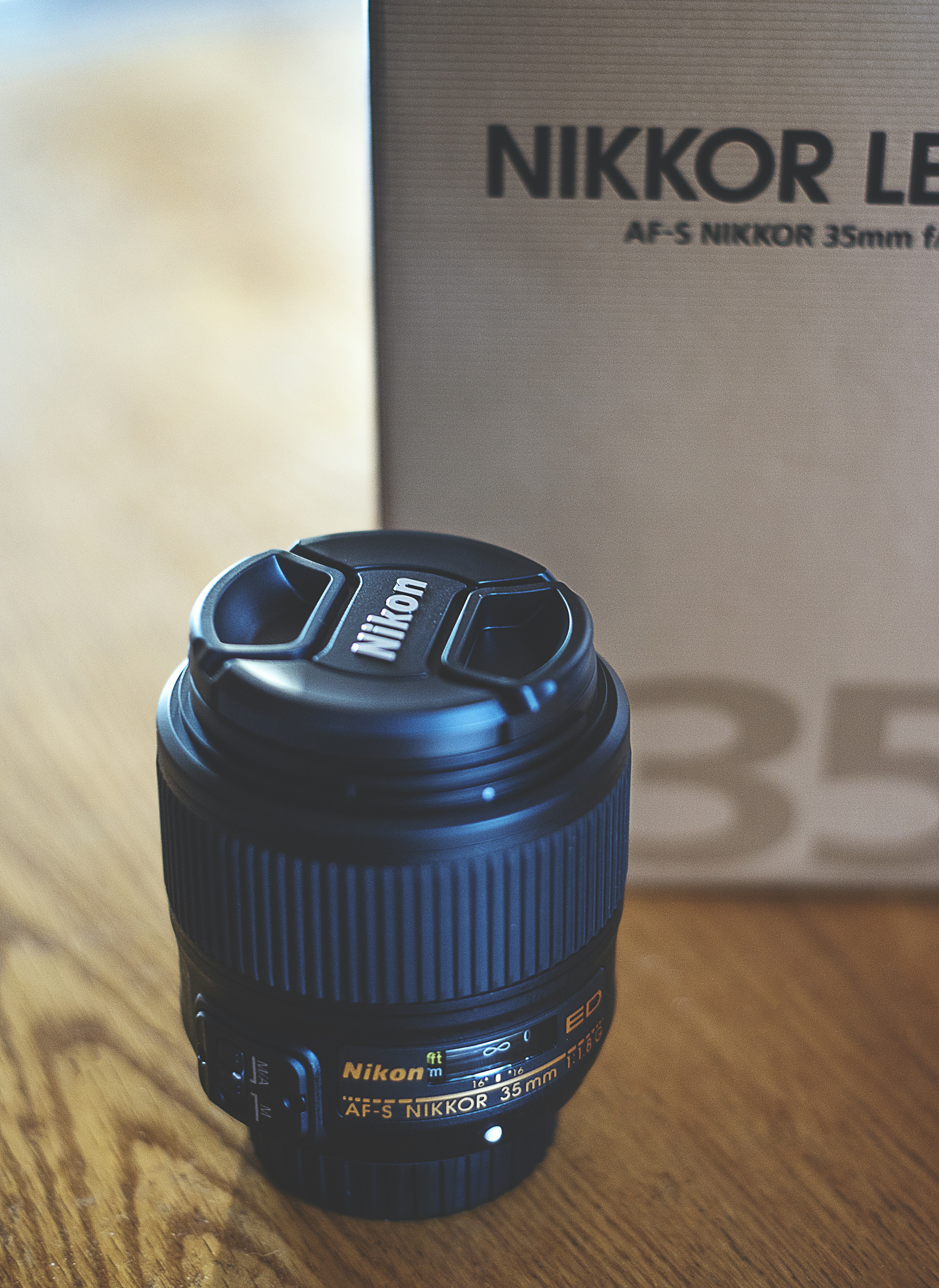 Close-up of a Nikon AF-S NIKKOR 35mm lens resting on a wooden surface with a blurred box in the background. The shot highlights the lens branding and compact form.