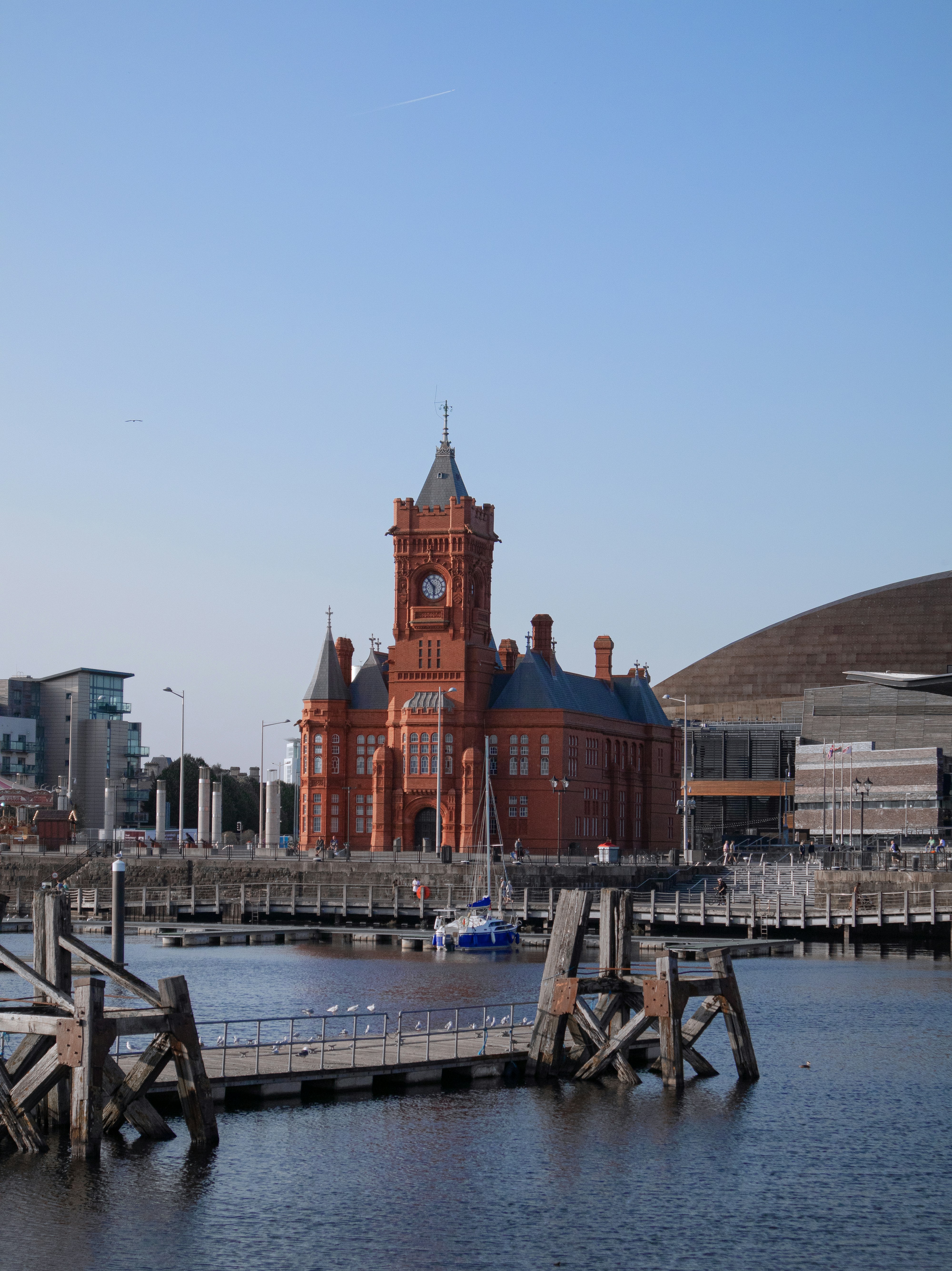 Historic red clocktower stands prominently by the waterfront, surrounded by modern architecture and wooden piers. A serene harbor scene unfolds under a clear blue sky.