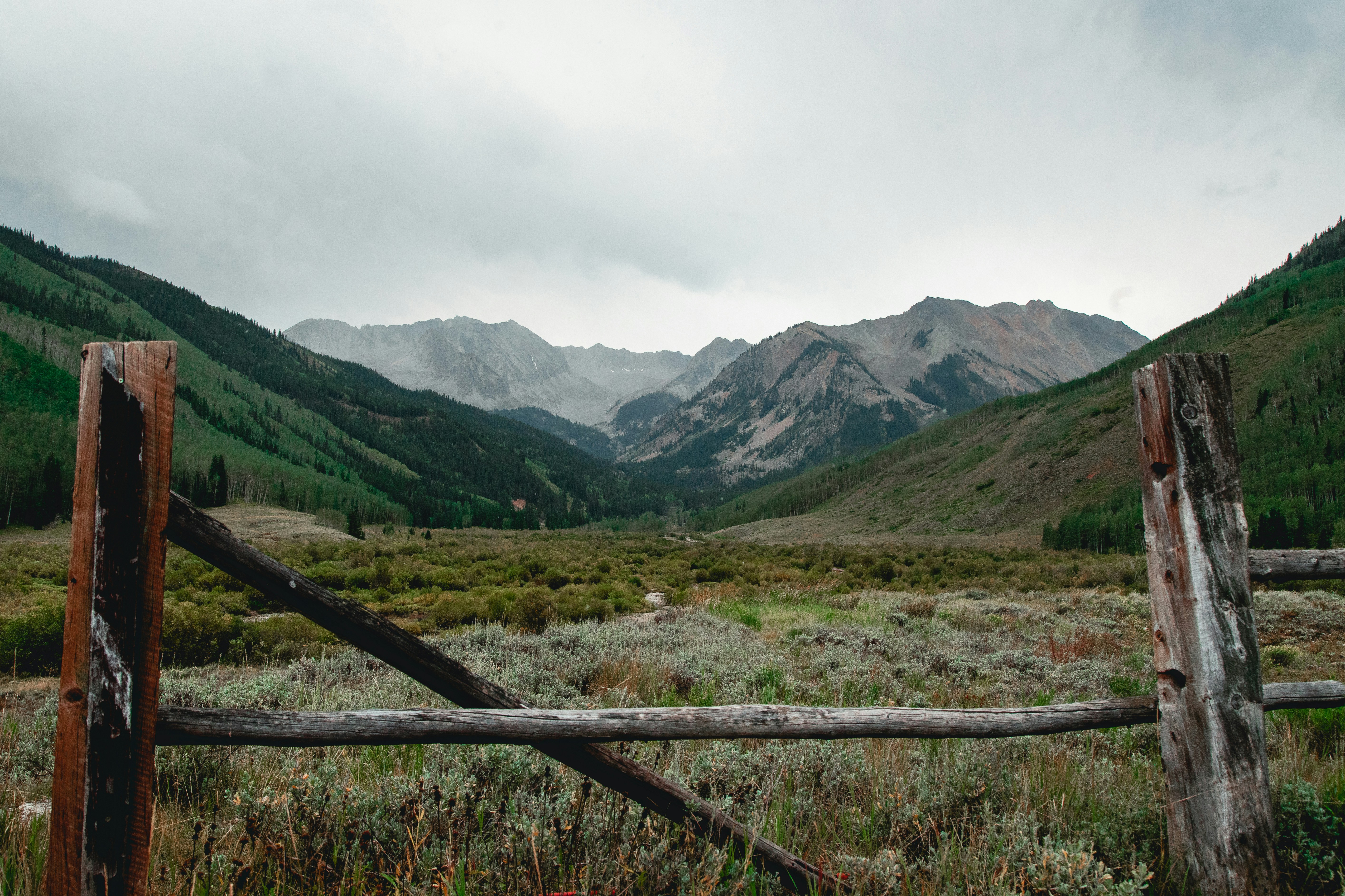 Rustic wooden fence frames a lush valley with distant mountain peaks under an overcast sky.