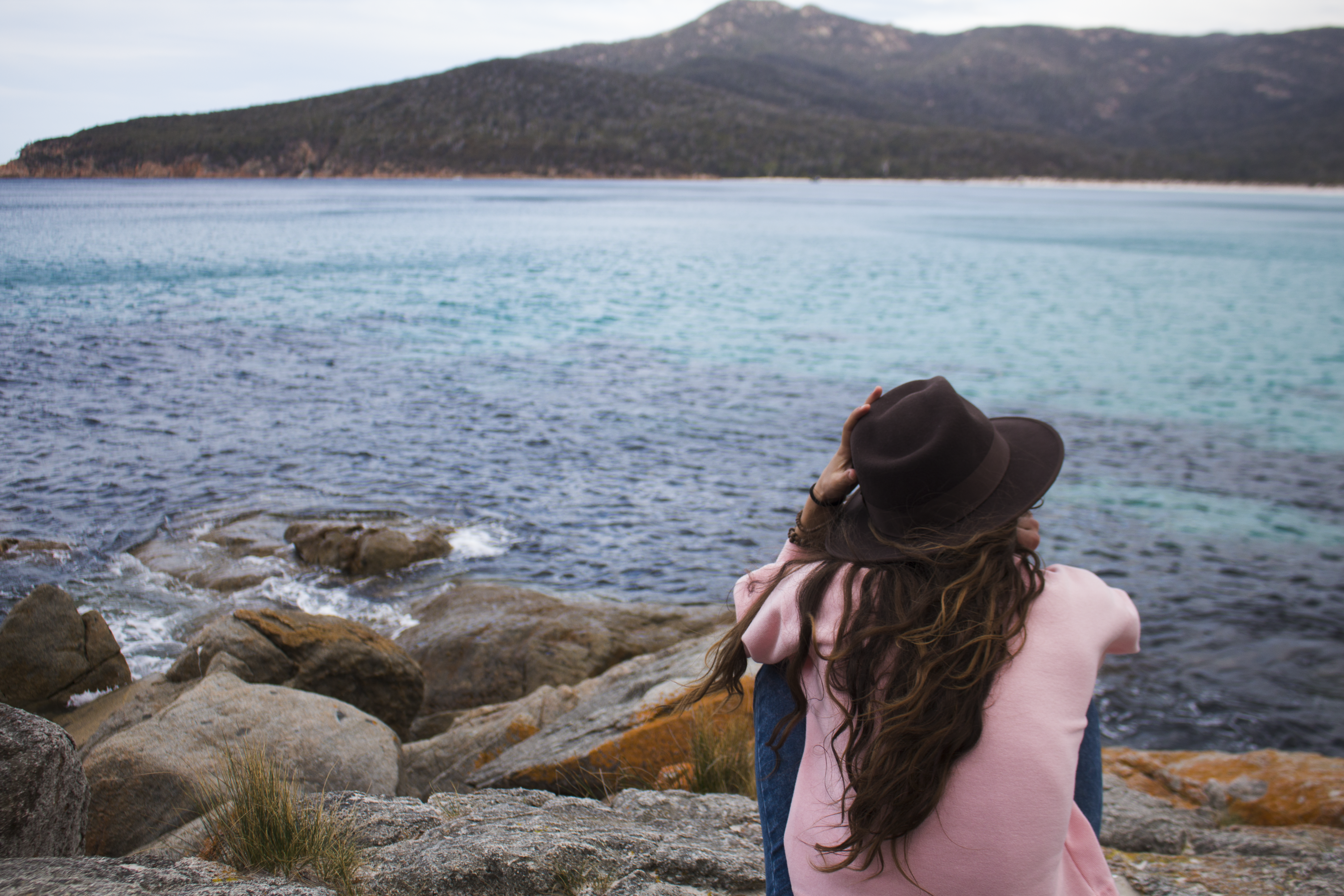 A serene scene captures a person sitting on rocky shores, gazing out at the vibrant blue ocean under an overcast sky. The composition highlights the contrast between the rugged rocks and the smooth water, with the person adding a contemplative mood. The colors are rich, with the sea's gradient from turquoise to deep blue creating a visually striking atmosphere, enhanced by the muted tones of the landscape.