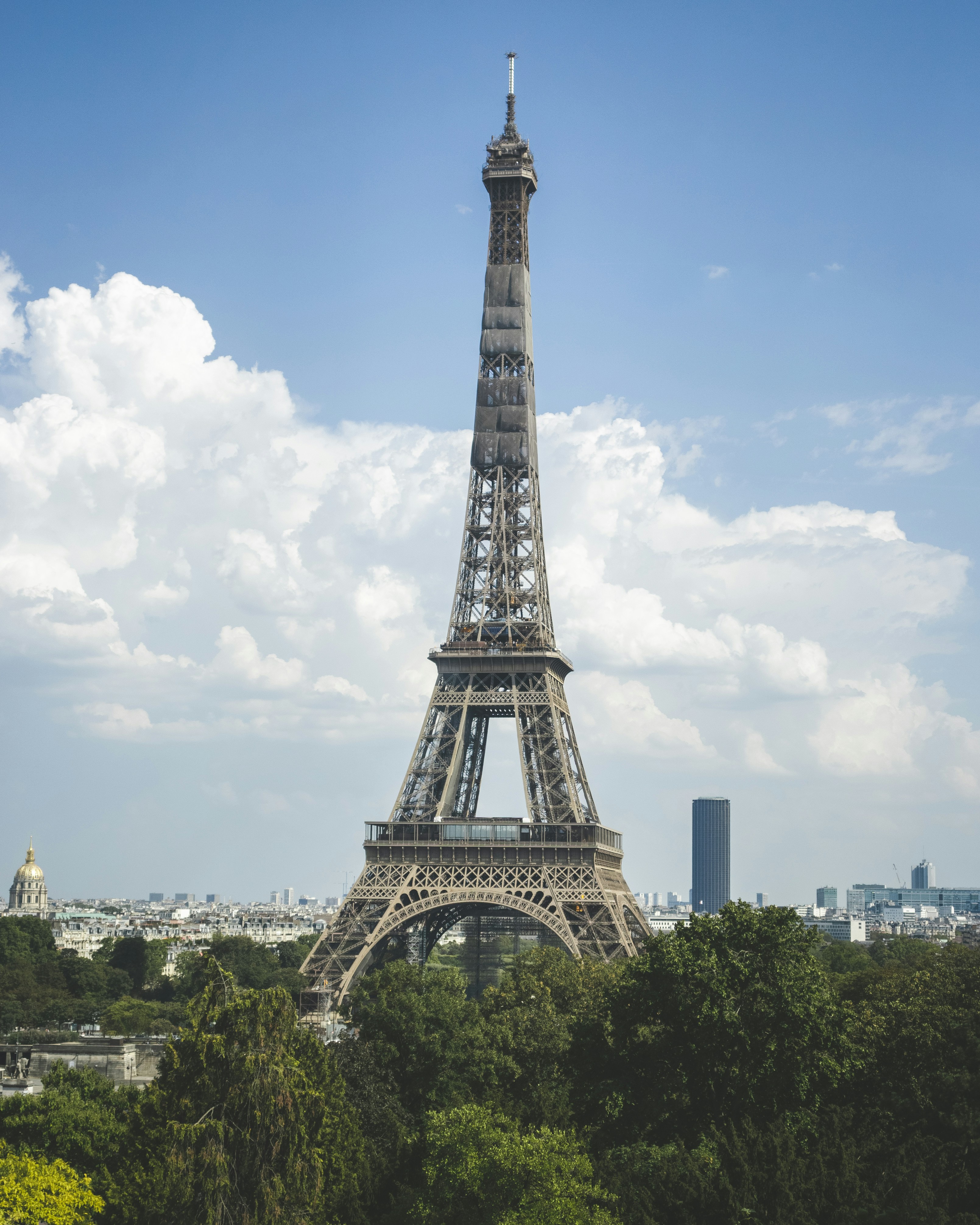 Eiffel Tower rises majestically above lush greenery and urban skyline under a blue sky dotted with clouds.
