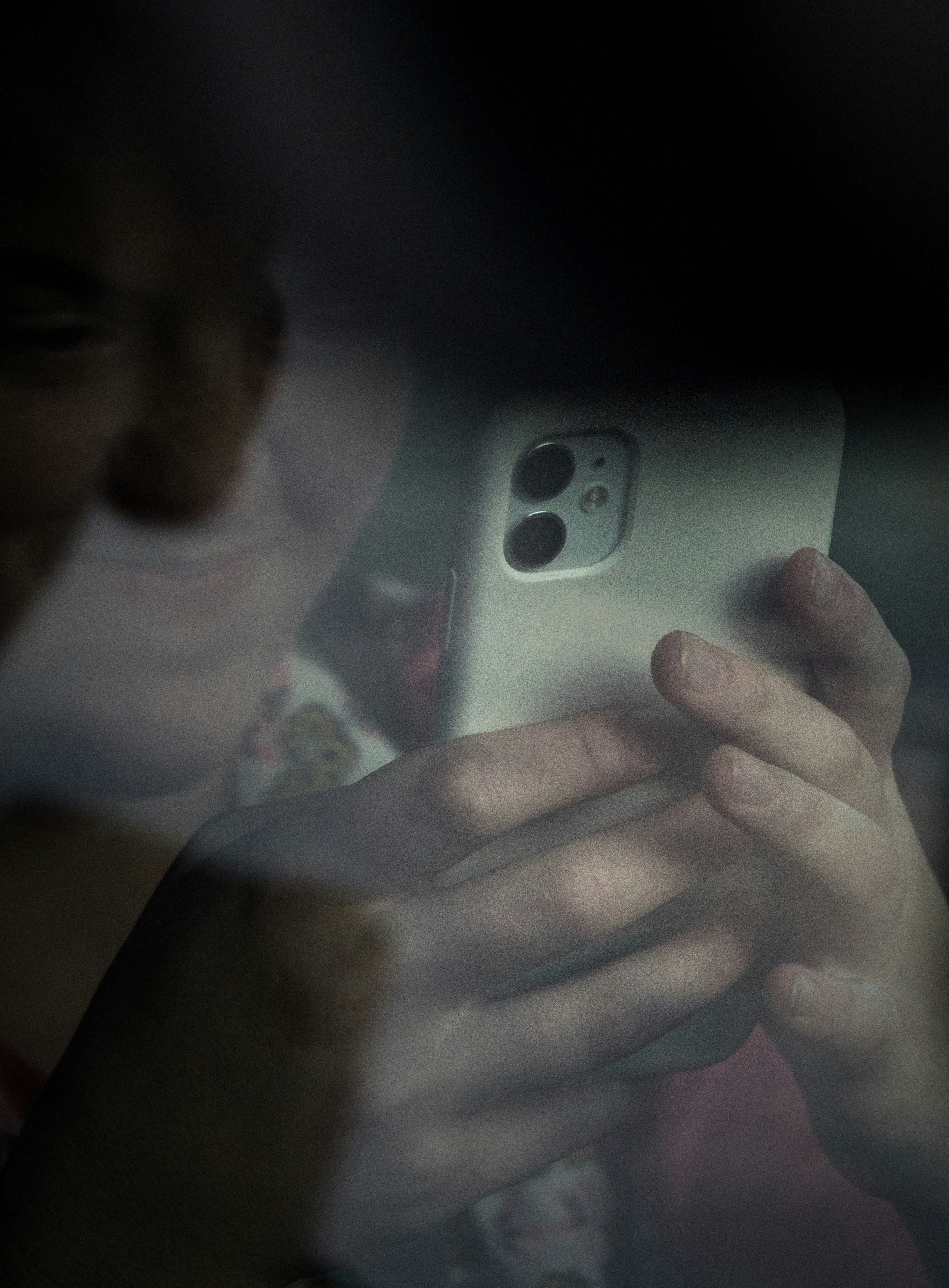 Dimly lit reflection of a person holding a silver iPhone behind a glass surface. The phone’s triple-camera module sits prominently in the frame.