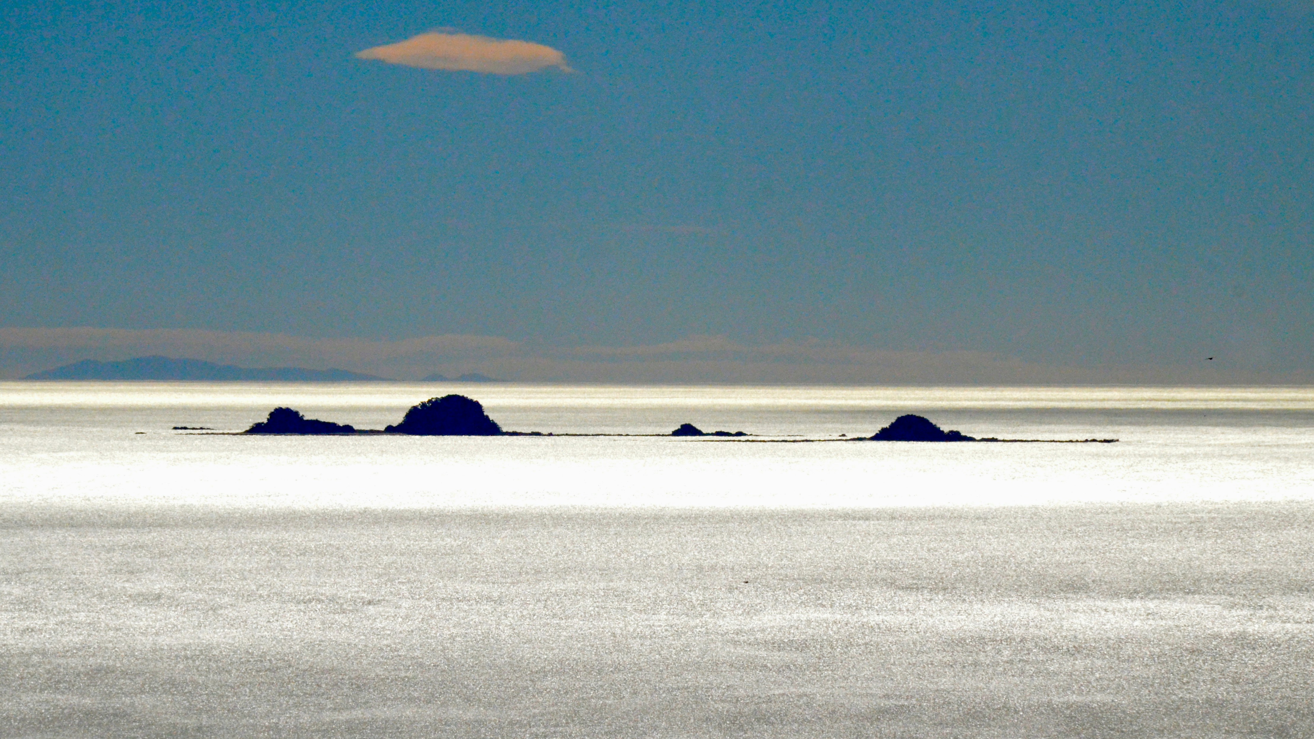 Distant islands rise from a shimmering sea under a deep blue sky with a lone cloud.