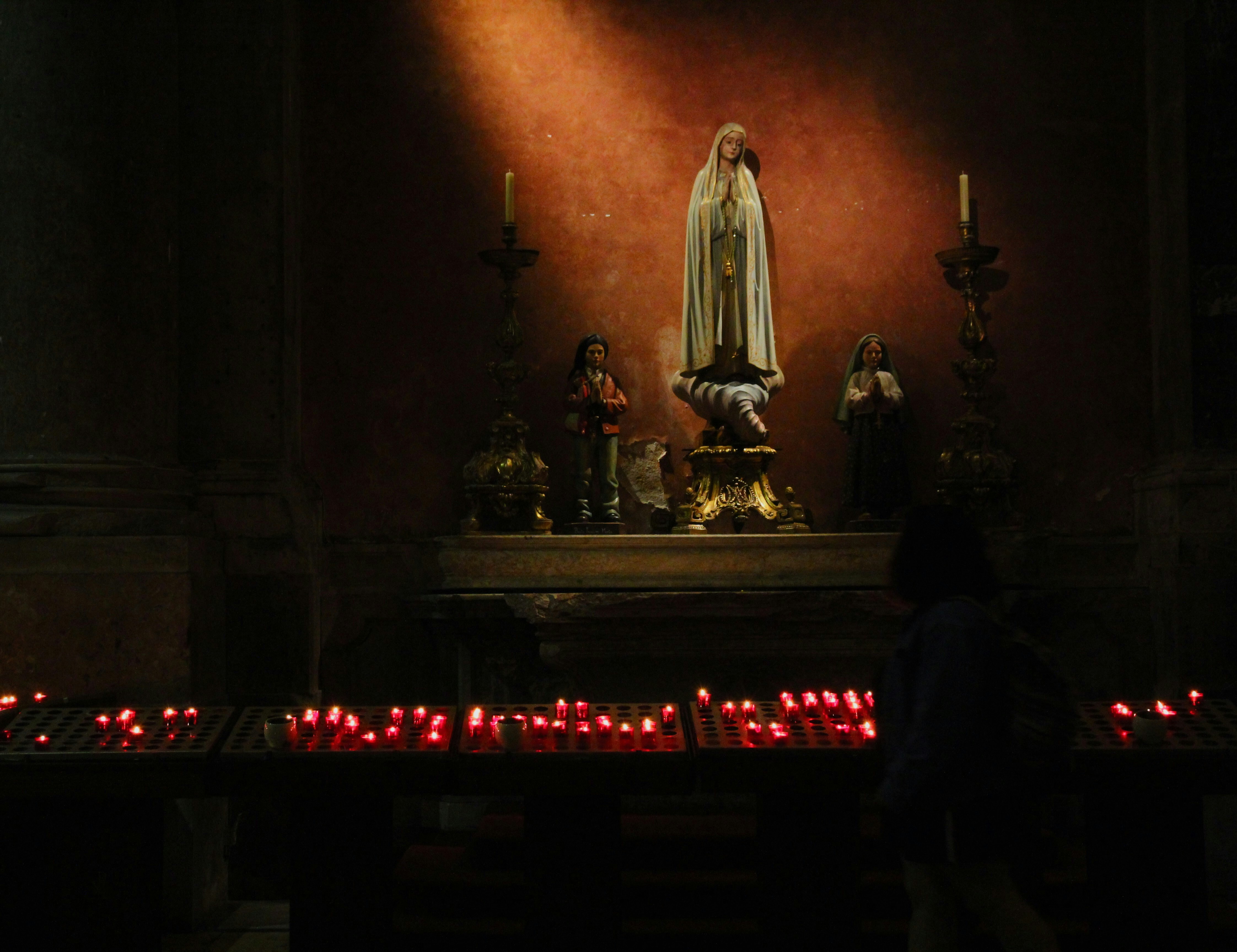 A serene altar scene featuring a statue of the Virgin Mary surrounded by lit candles, evoking a sense of peace and contemplation.