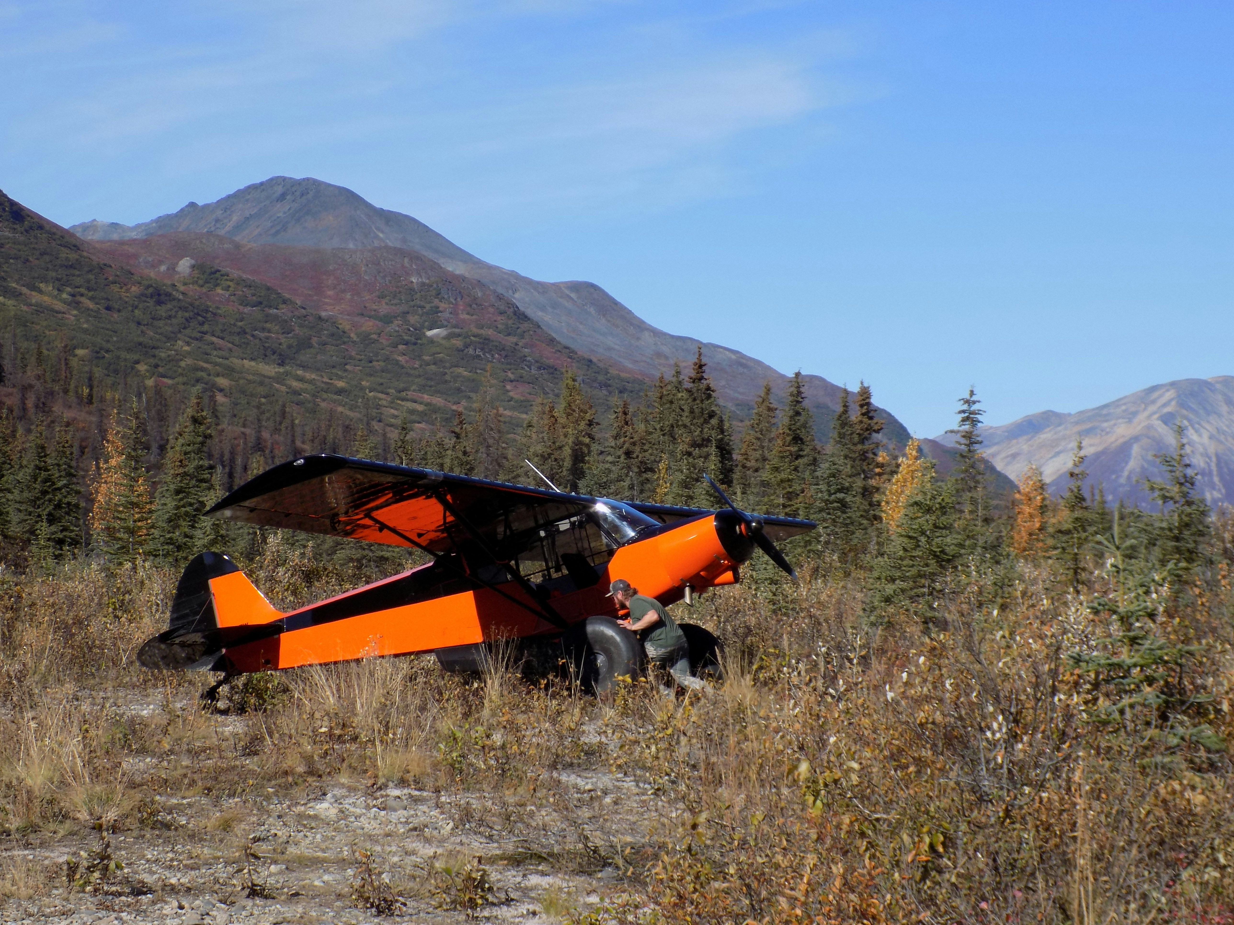 An orange bush plane parked amidst the rugged terrain of the Alaskan wilderness, surrounded by autumn foliage and mountains in the background.