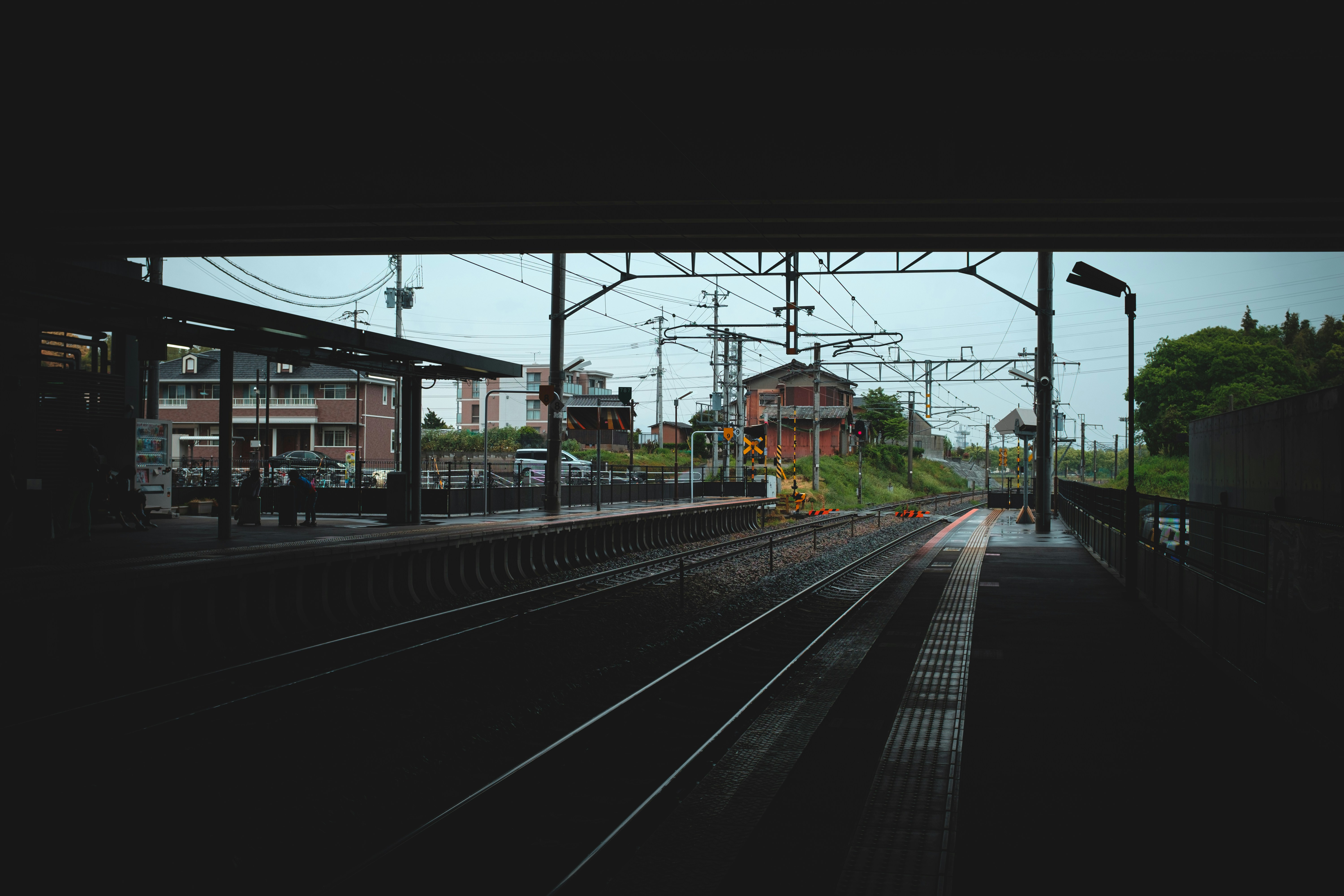 Dimly lit train station platform with visible tracks leading into the distance, framed by a canopy and surrounding buildings.
