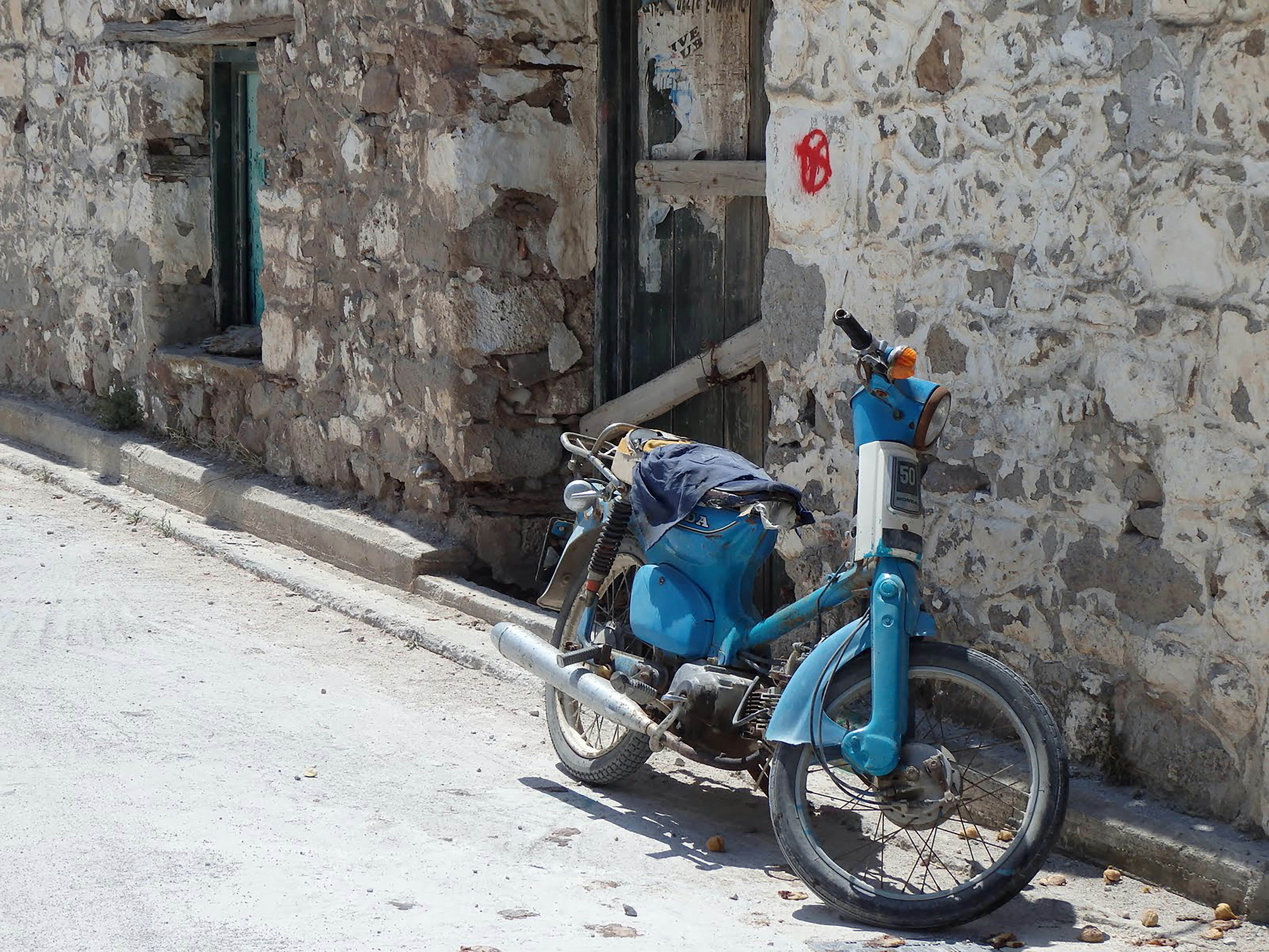 Blue vintage-style scooter parked along a sunlit stone wall on a narrow street. Photograph capturing a quiet urban moment with the bike as the focal subject.