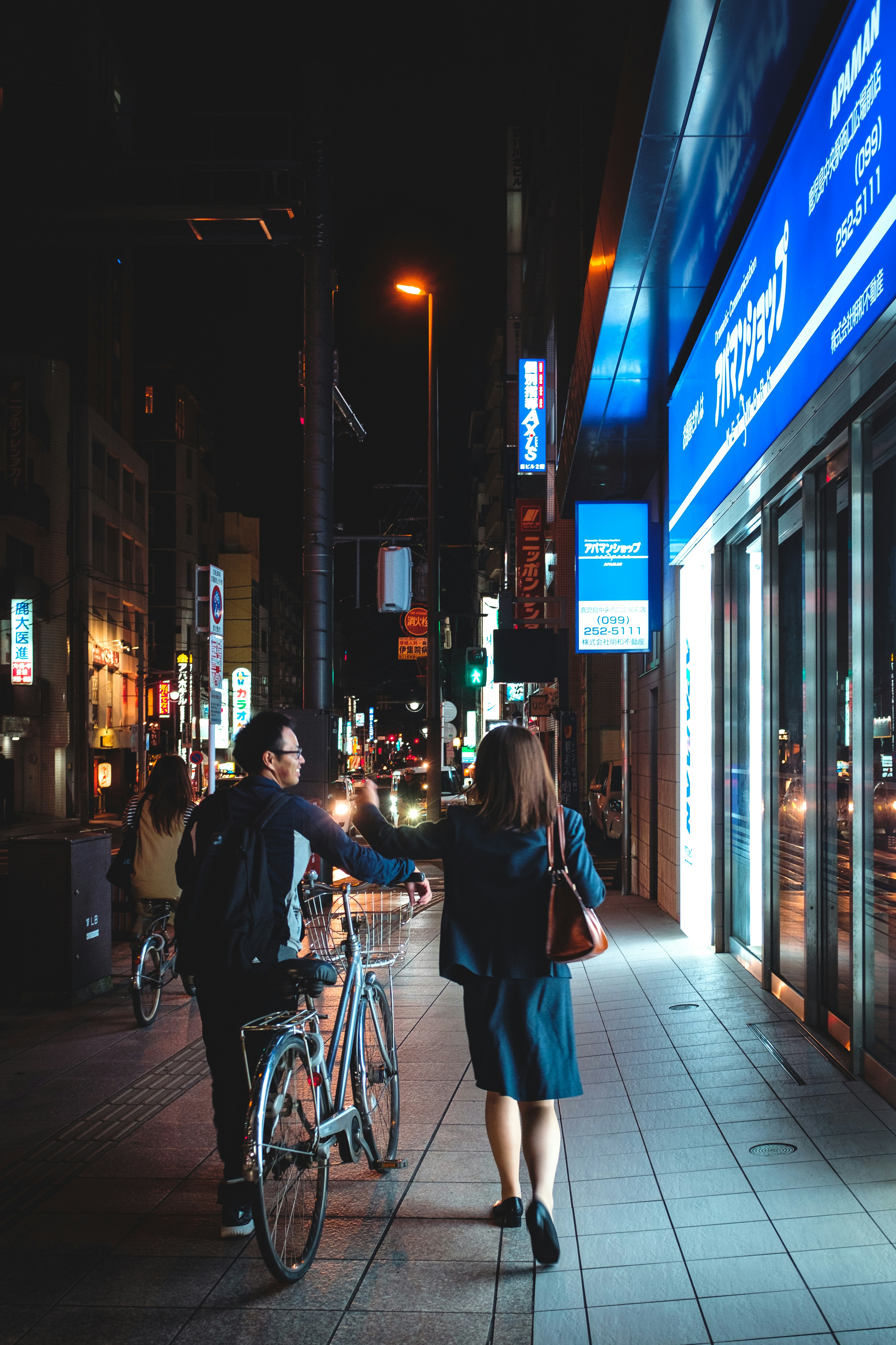 Two pedestrians share a moment on a bustling city street illuminated by vibrant neon signs and storefronts.