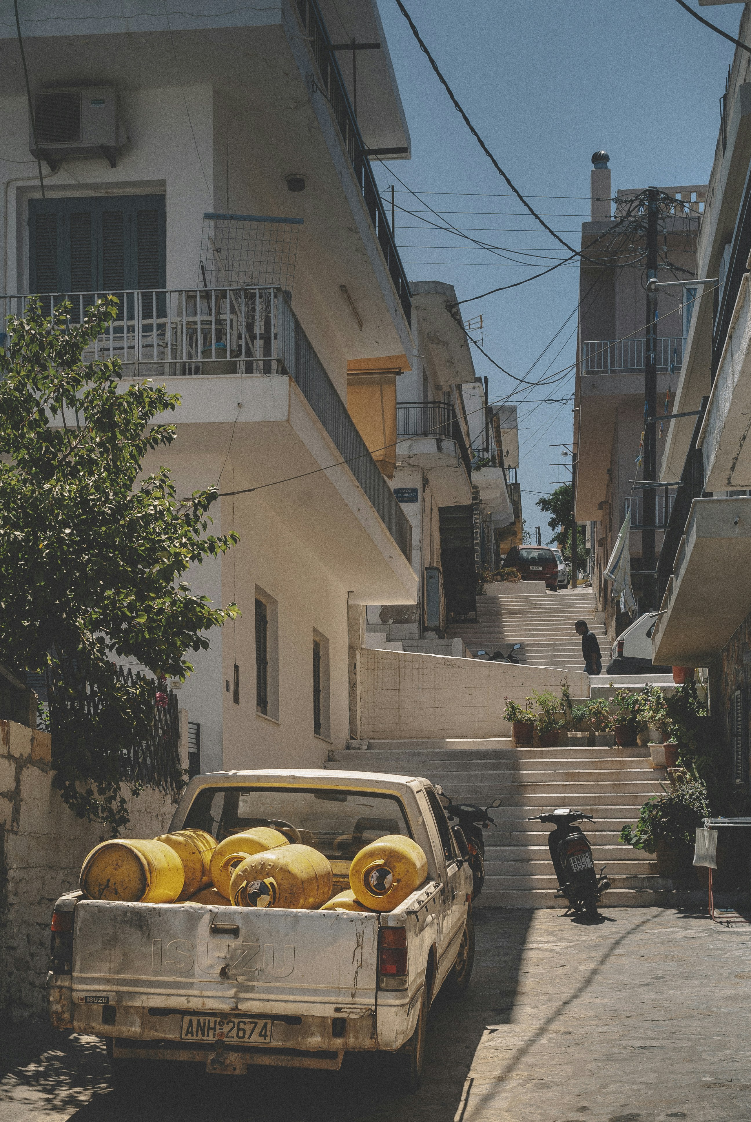 Narrow street lined with residential buildings and a parked truck filled with yellow barrels, leading up to a staircase adorned with potted plants.