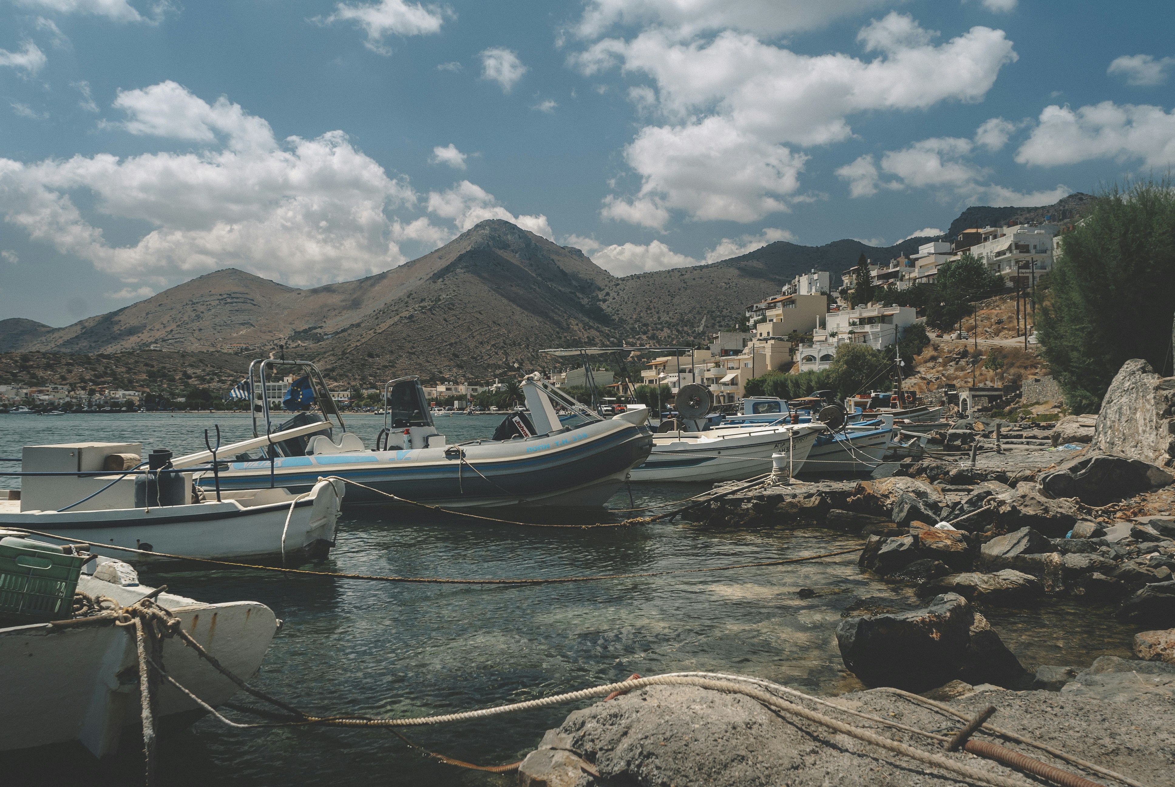 Fishing boats moored along a rocky shore with a backdrop of distant mountains and scattered clouds. The scene captures the tranquil essence of coastal living.