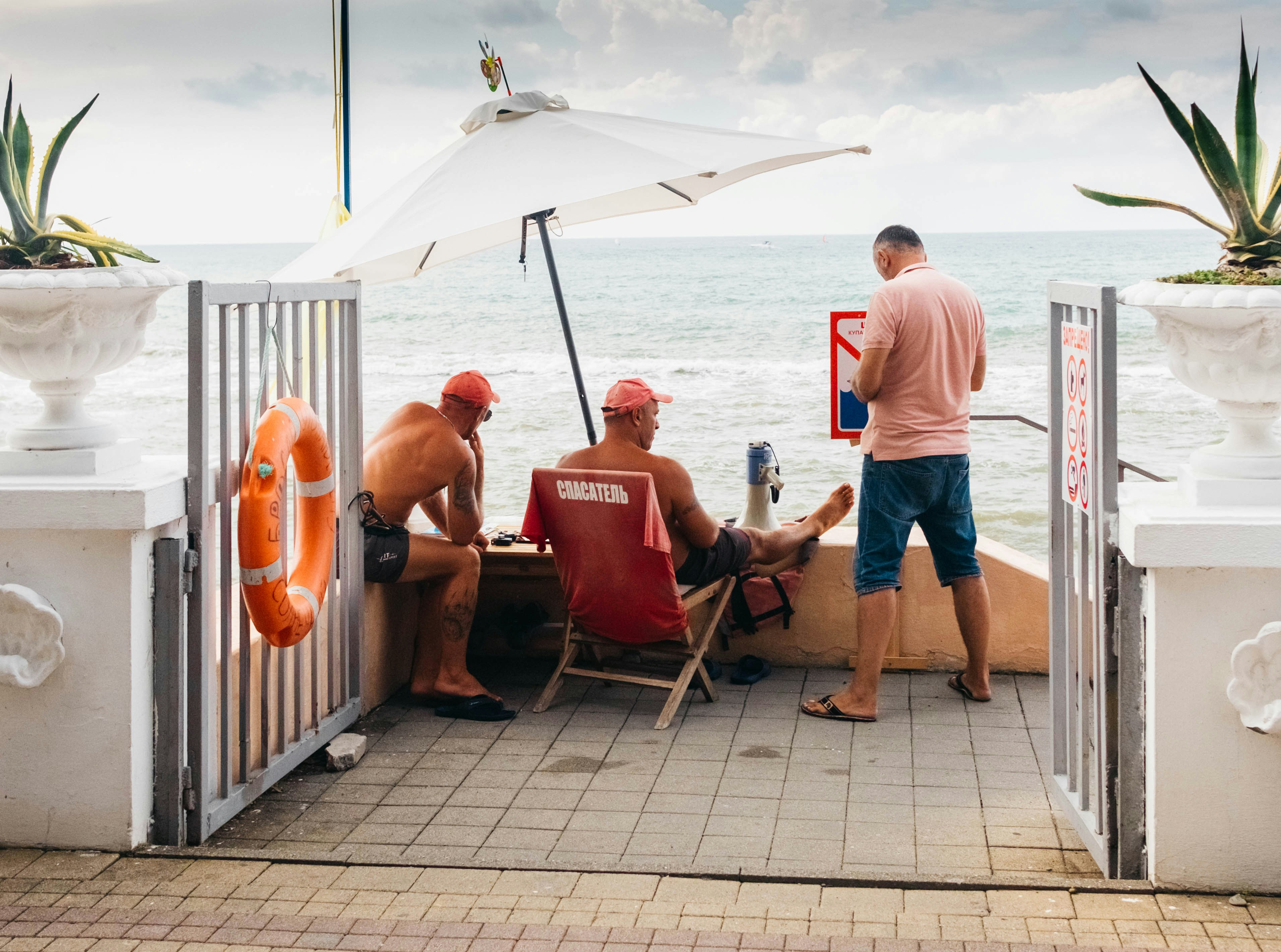 Three individuals relax by the ocean, with one seated in a lifeguard chair under an umbrella. A life buoy and warning signs enhance the seaside ambiance.