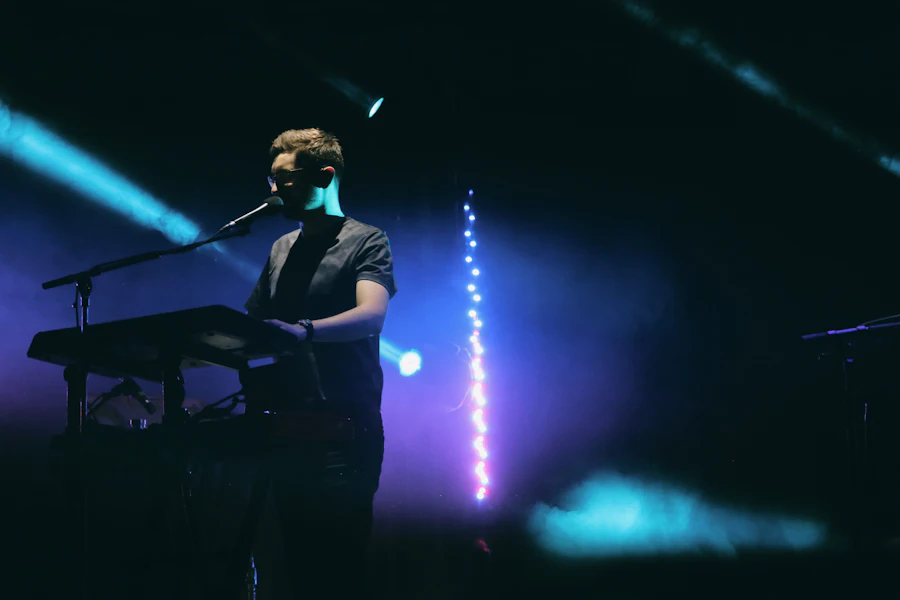 A musician stands at a keyboard and microphone on a dark concert stage, lit by blue and purple beams with haze in the background.