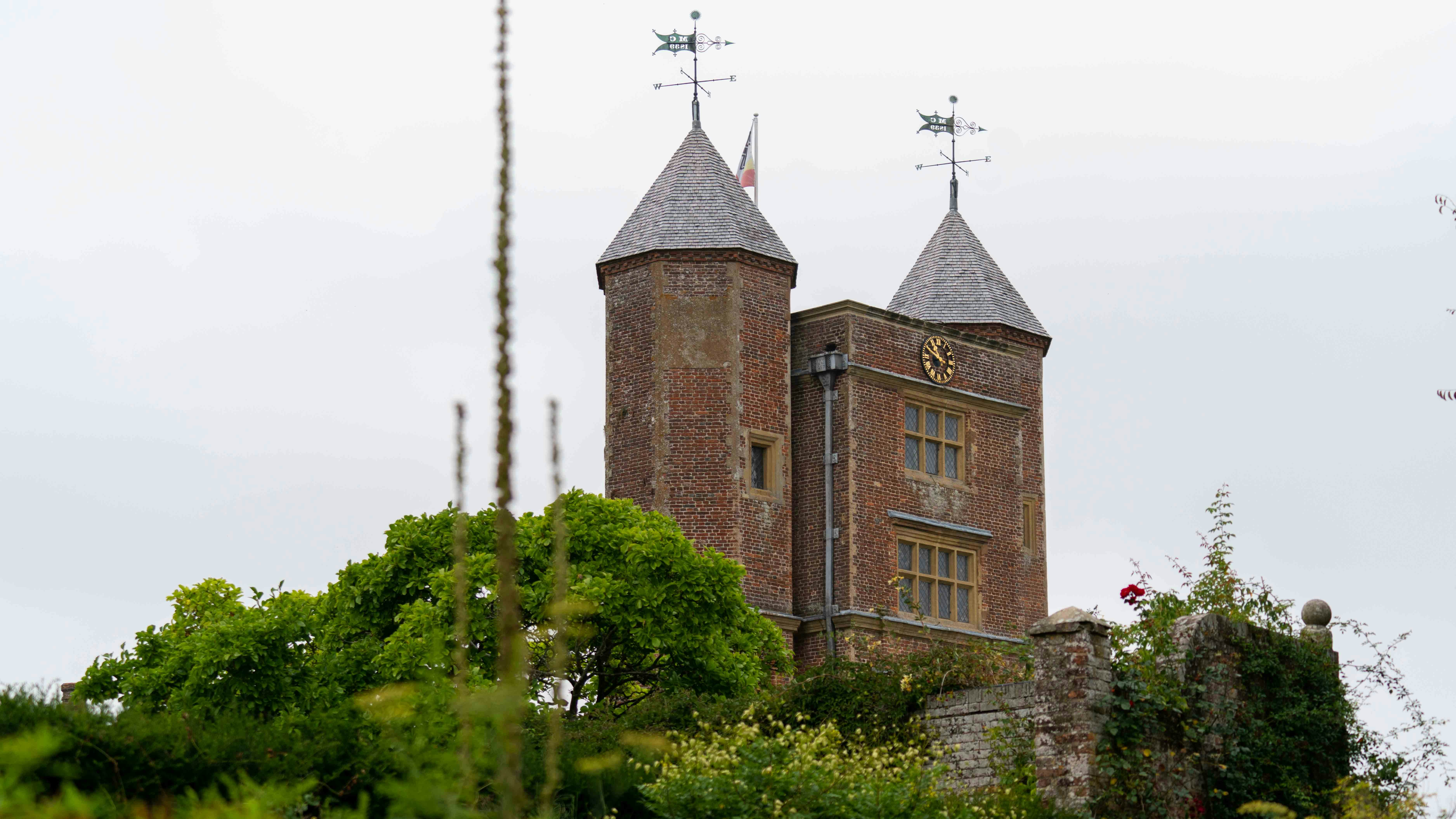 Historic tower with distinctive weather vanes and a clock, surrounded by lush greenery and a stone wall.