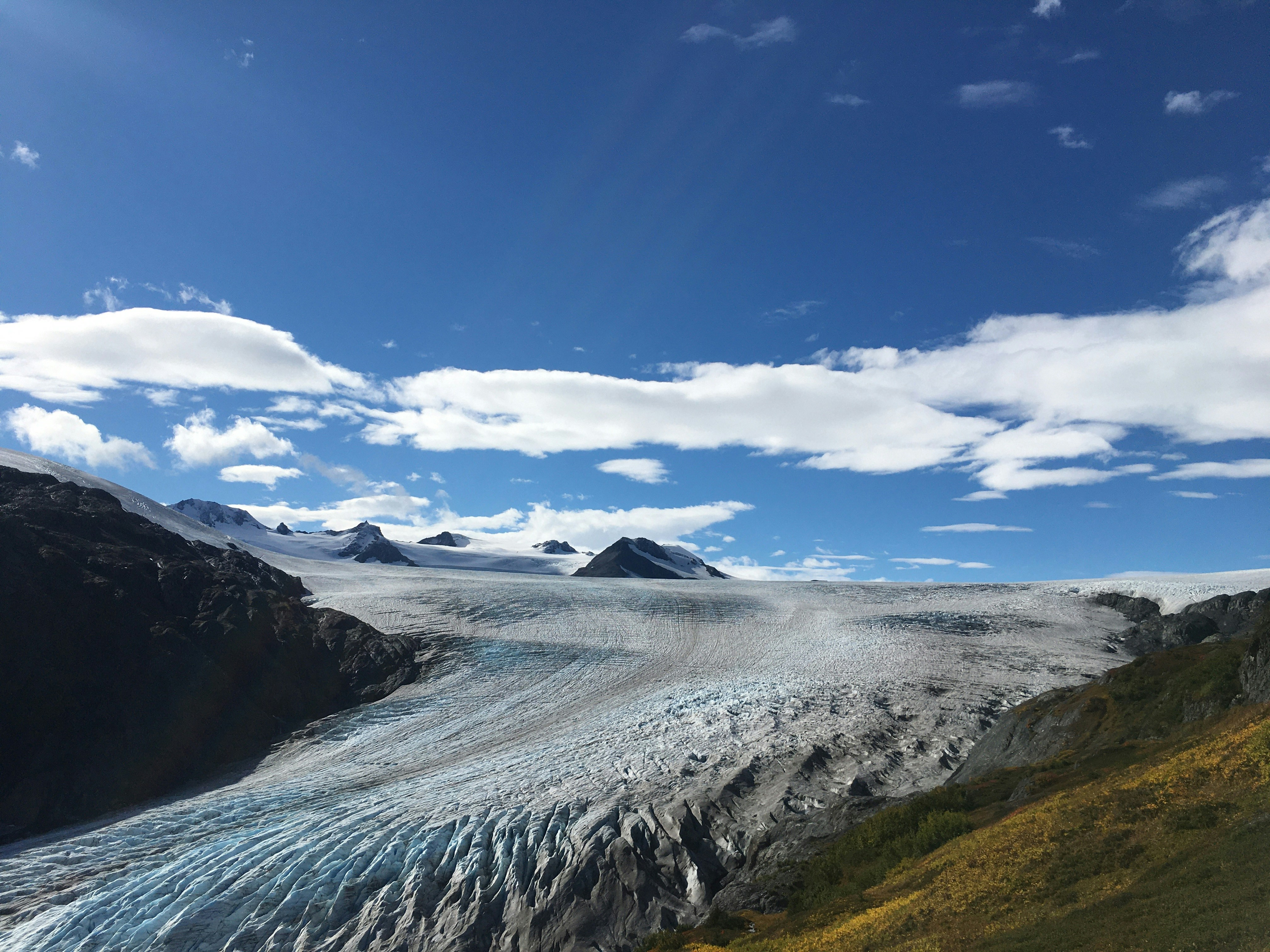 Expansive glacier flows beneath a bright blue sky dotted with clouds, framed by rugged mountains in the distance. A hint of autumn foliage adds a splash of color to the scene.