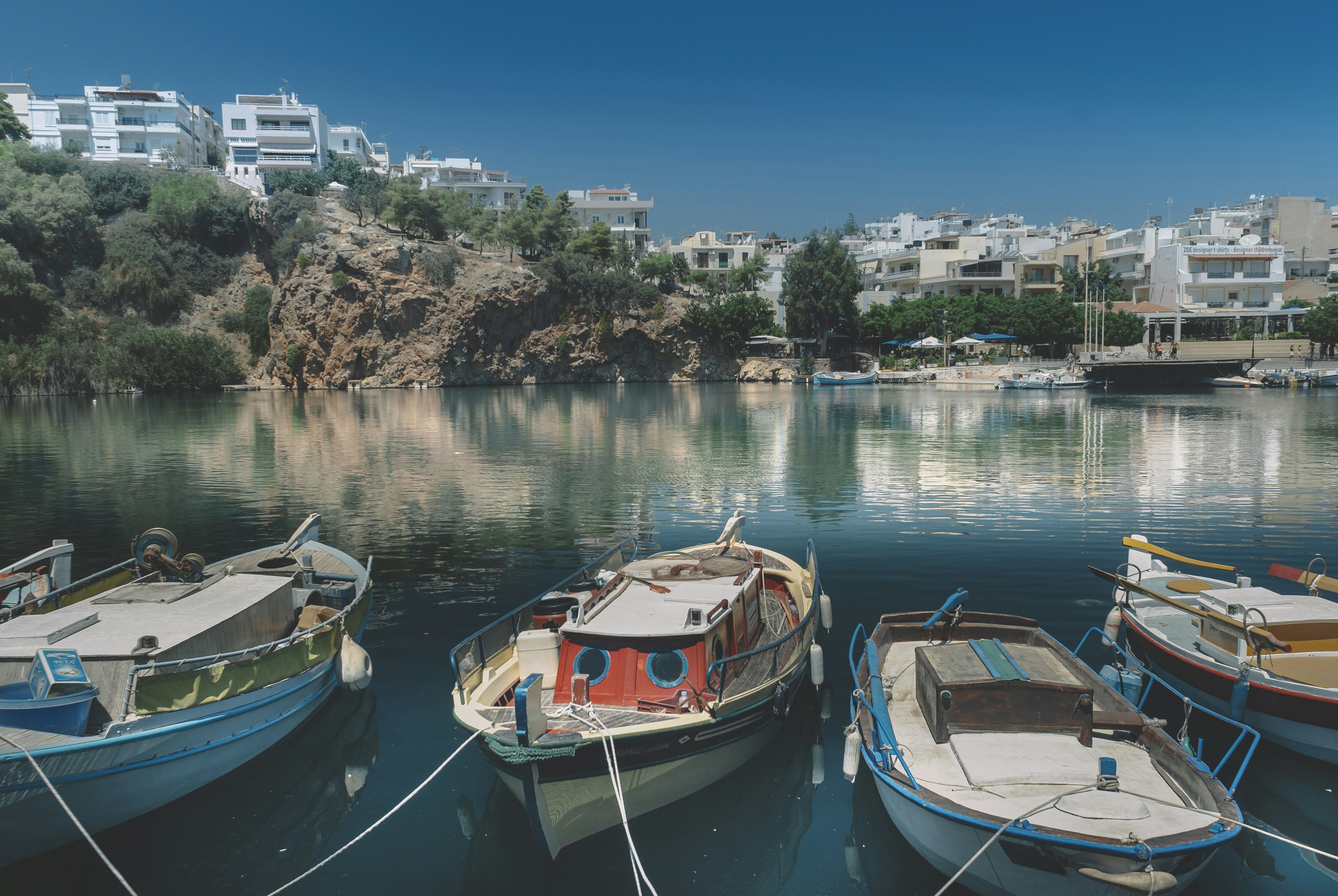 Colorful fishing boats moored in a tranquil harbor, surrounded by modern buildings and lush greenery reflecting in the calm waters.