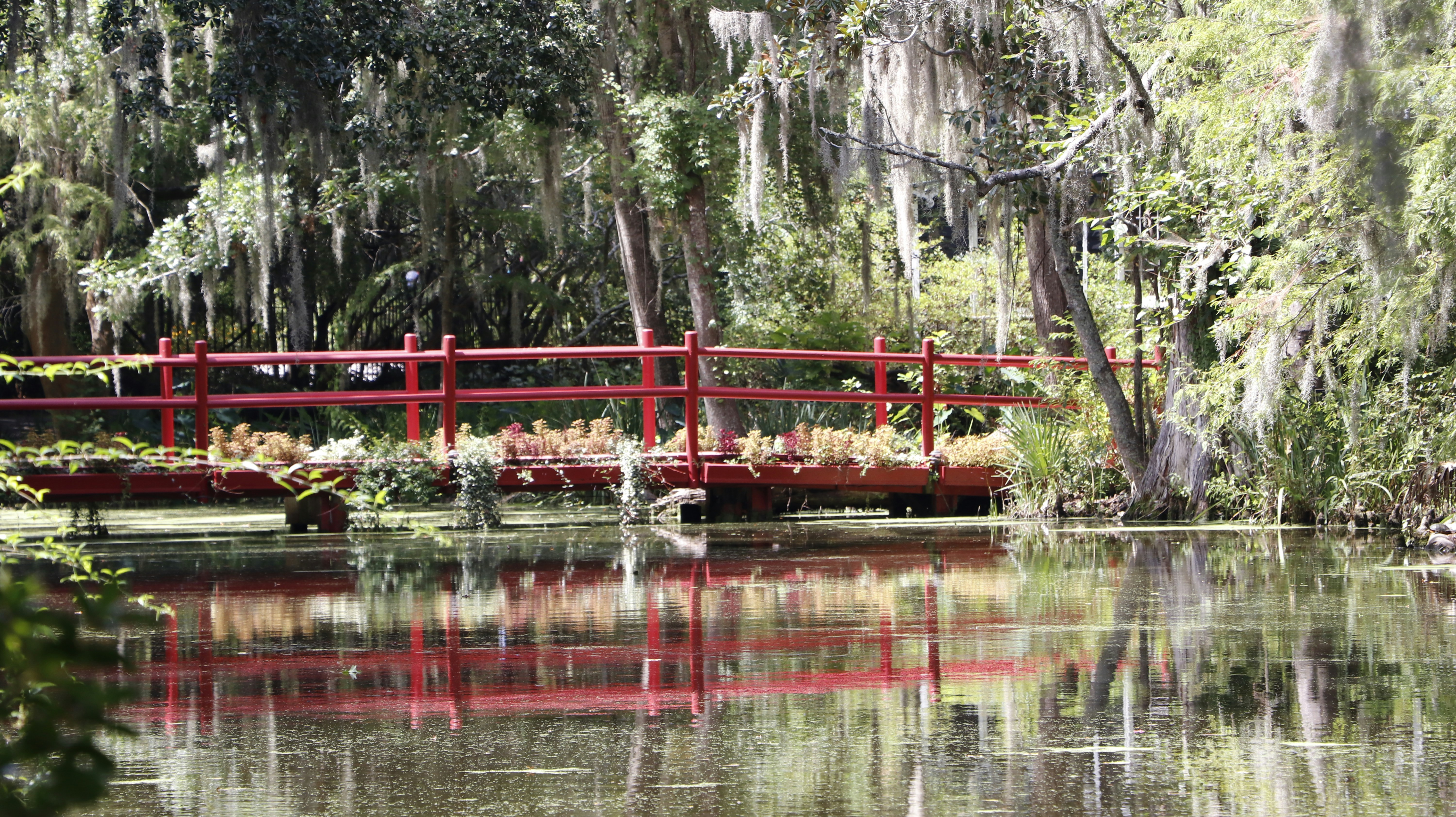 Red bridge arches over a tranquil river, surrounded by lush greenery and reflected in the calm water.