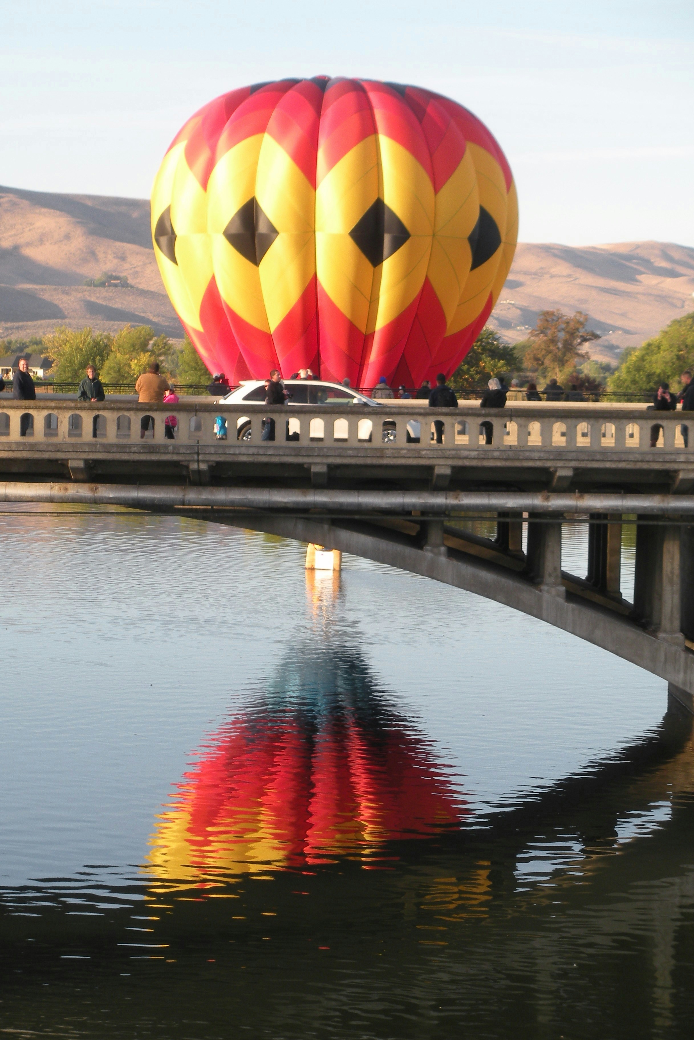 Red-yellow diamond-pattern hot air balloon hovers above a stone-arched bridge, its reflection shimmering in the calm river.