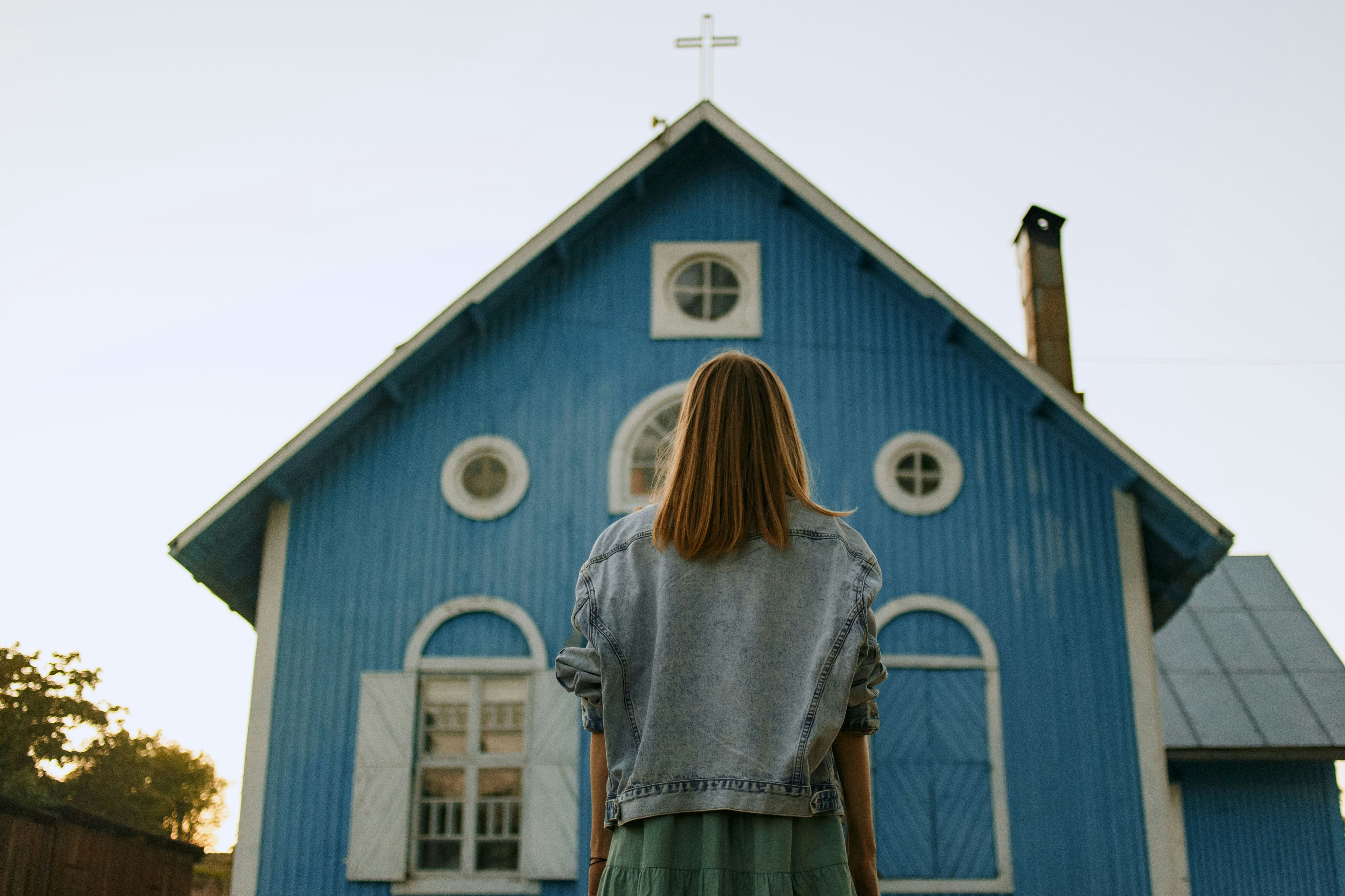 Person standing in front of a blue wooden chapel with arched windows and a cross on the roof.