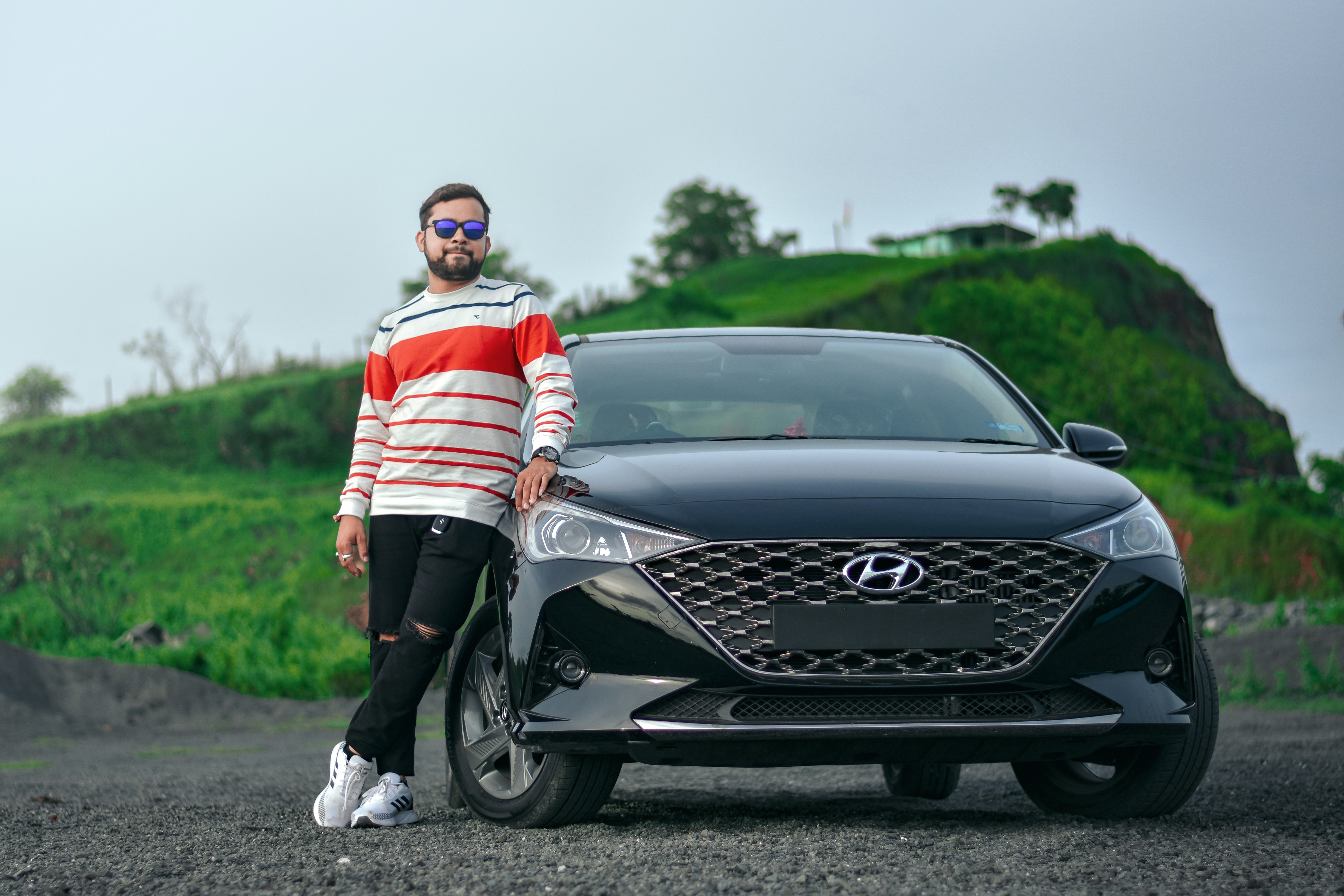 Man in stylish attire leans against a sleek black Hyundai car with a lush green hill in the background. The scene conveys a modern lifestyle and automotive elegance.