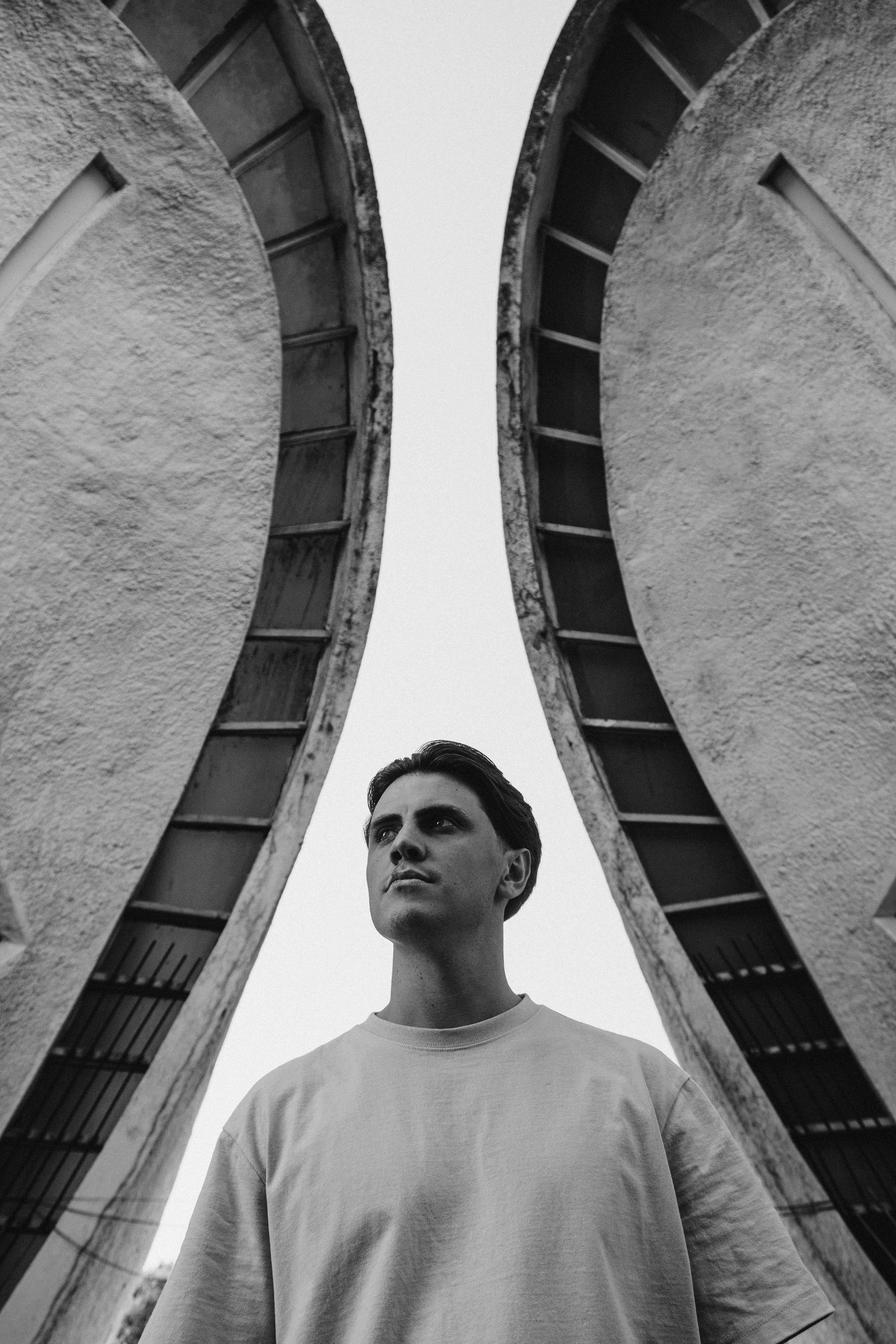 A young man stands thoughtfully between two towering architectural structures, captured in striking black and white. The symmetry and lines create a dramatic visual effect.