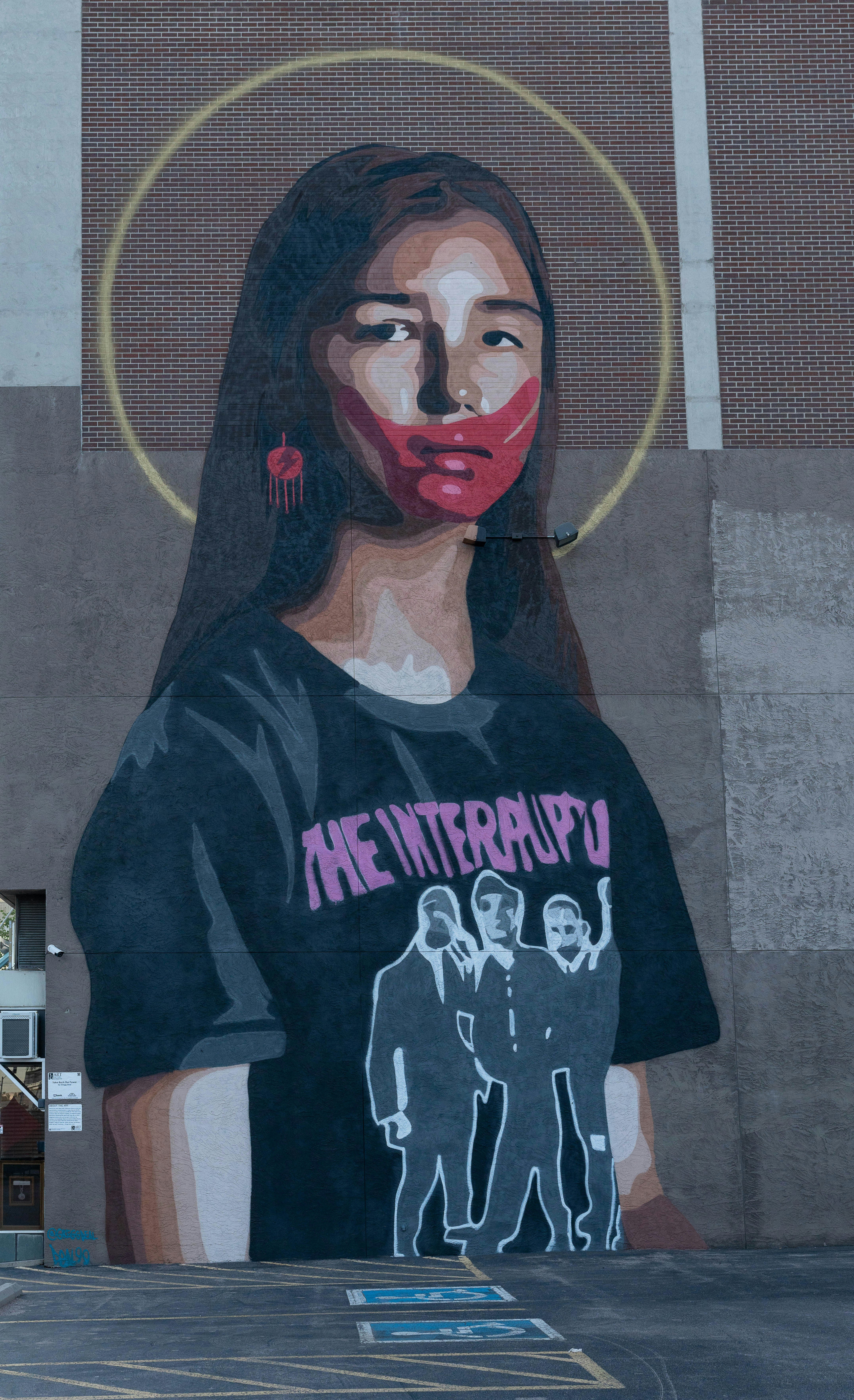 Vibrant mural depicting a young woman with a halo, wearing a shirt that reads 'THE INTERRUPT.' The artwork emphasizes cultural identity and empowerment.