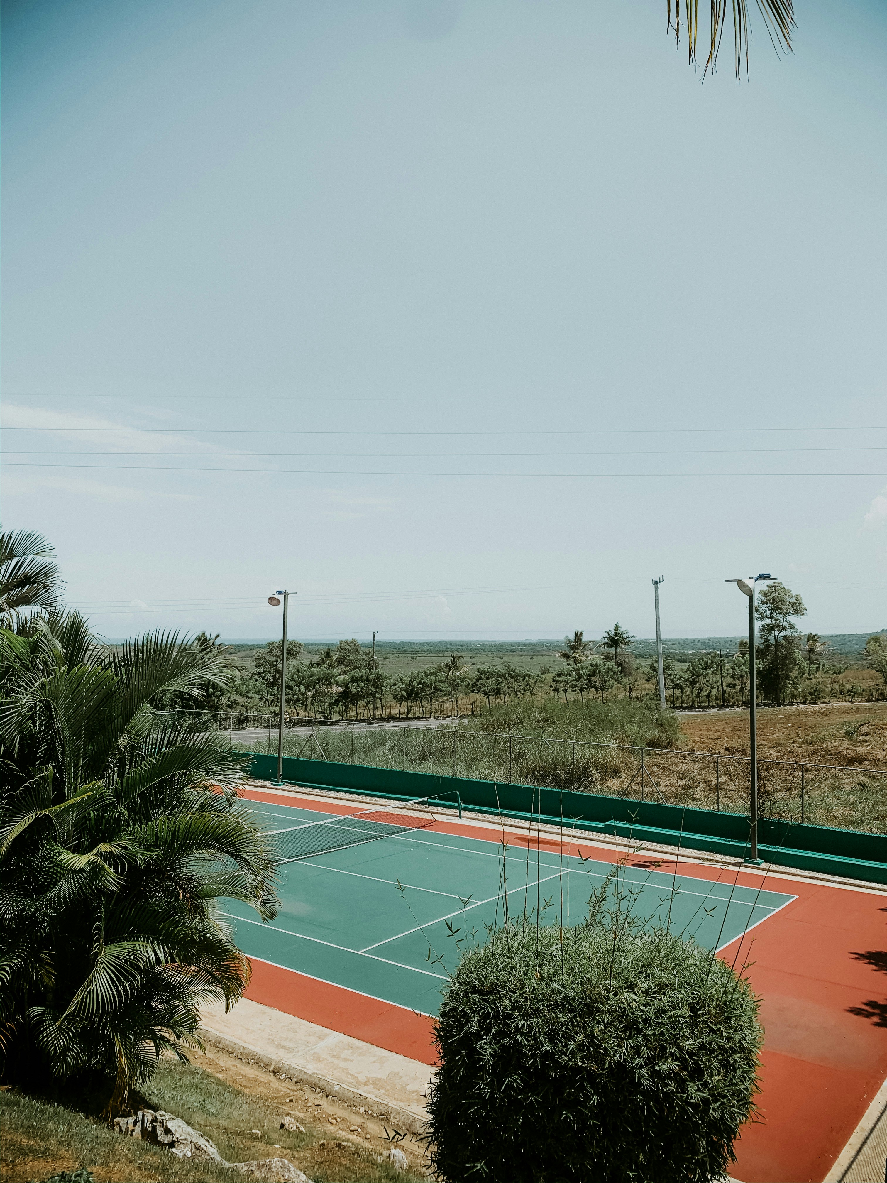 Tennis court framed by lush greenery, set against a backdrop of expansive fields and a clear sky. The scene conveys tranquility and sport.