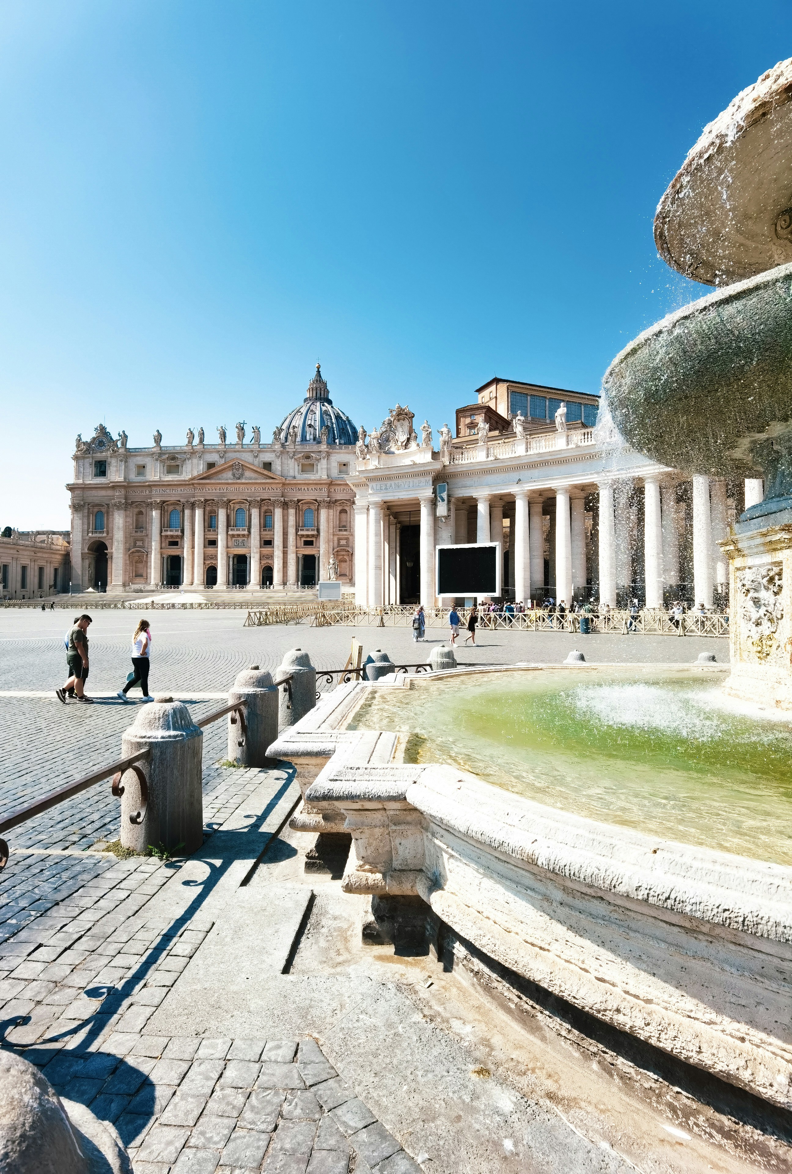 Sunlit Vatican Piazza featuring the dome of St. Peter's Basilica and the colonnade, with a multi-tier fountain in the foreground and a few pedestrians.