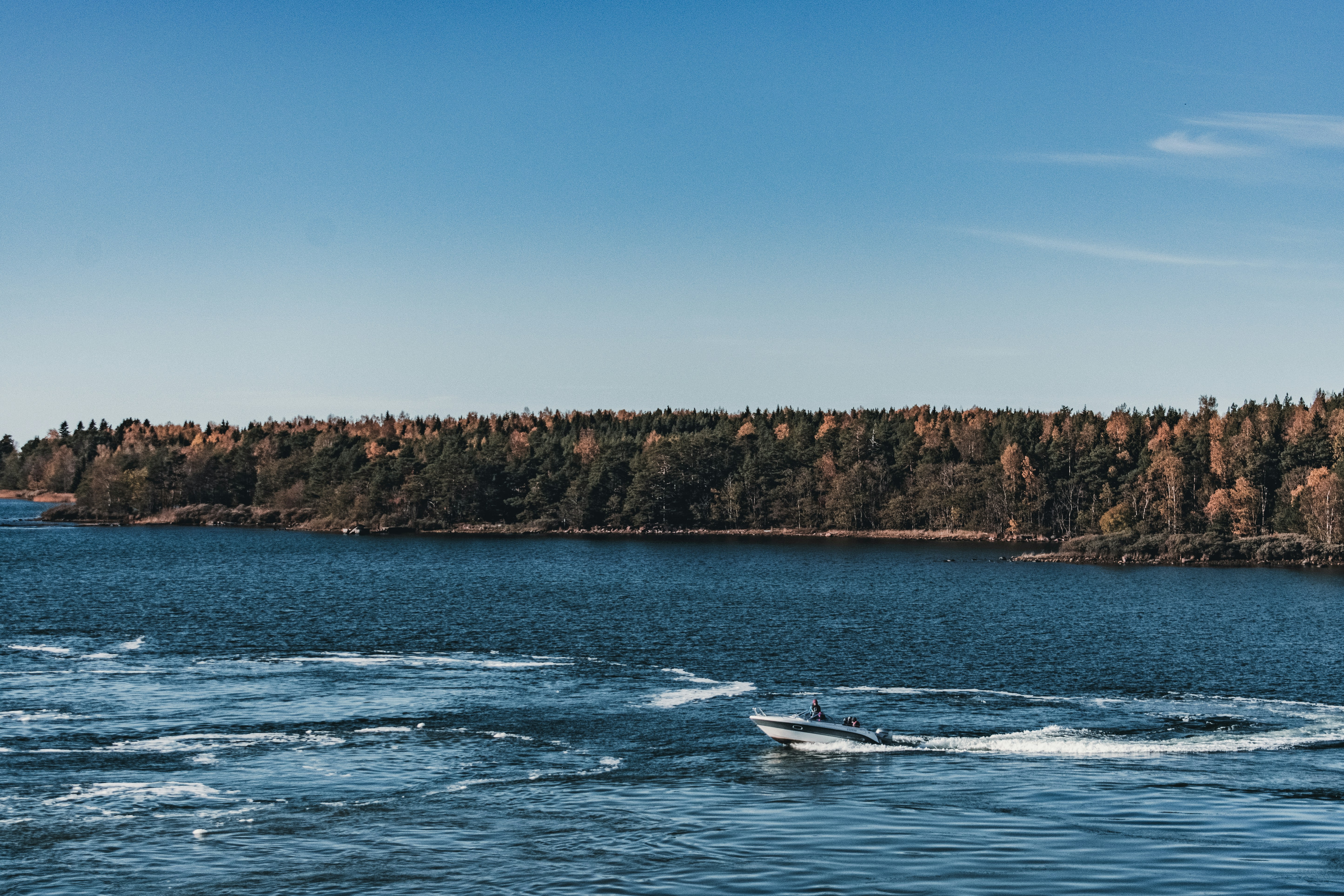 A speedboat glides through calm waters, leaving a trail behind, with a backdrop of autumn-colored trees lining the shore.