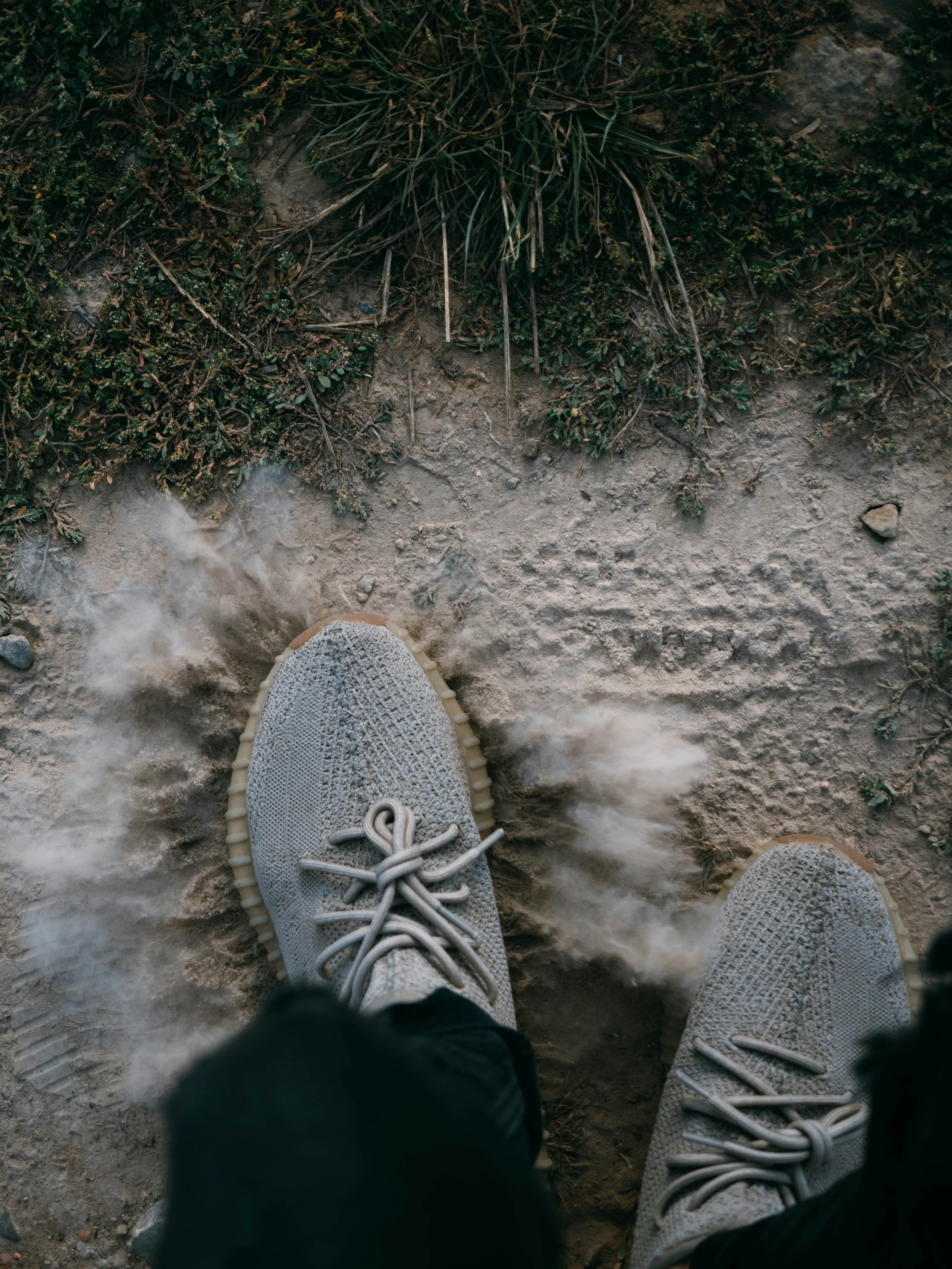 A pair of gray sneakers leaving a trail of dust on a dirt path, surrounded by grass. The scene captures a moment of exploration.