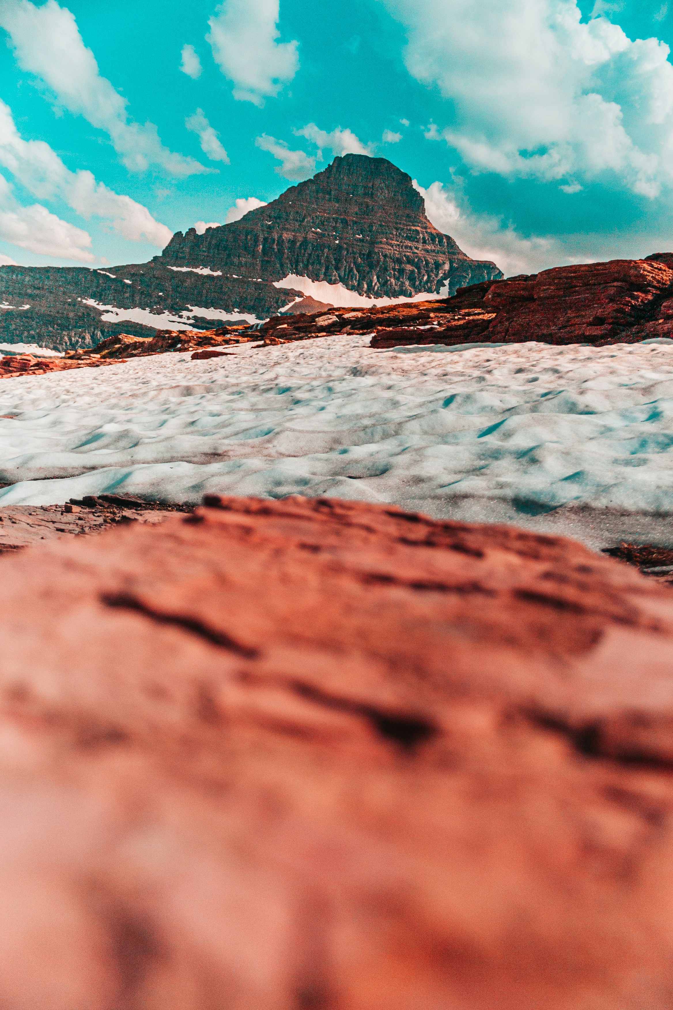 Rugged red rock formations lead to a towering mountain, framed by a bright blue sky dotted with fluffy clouds.