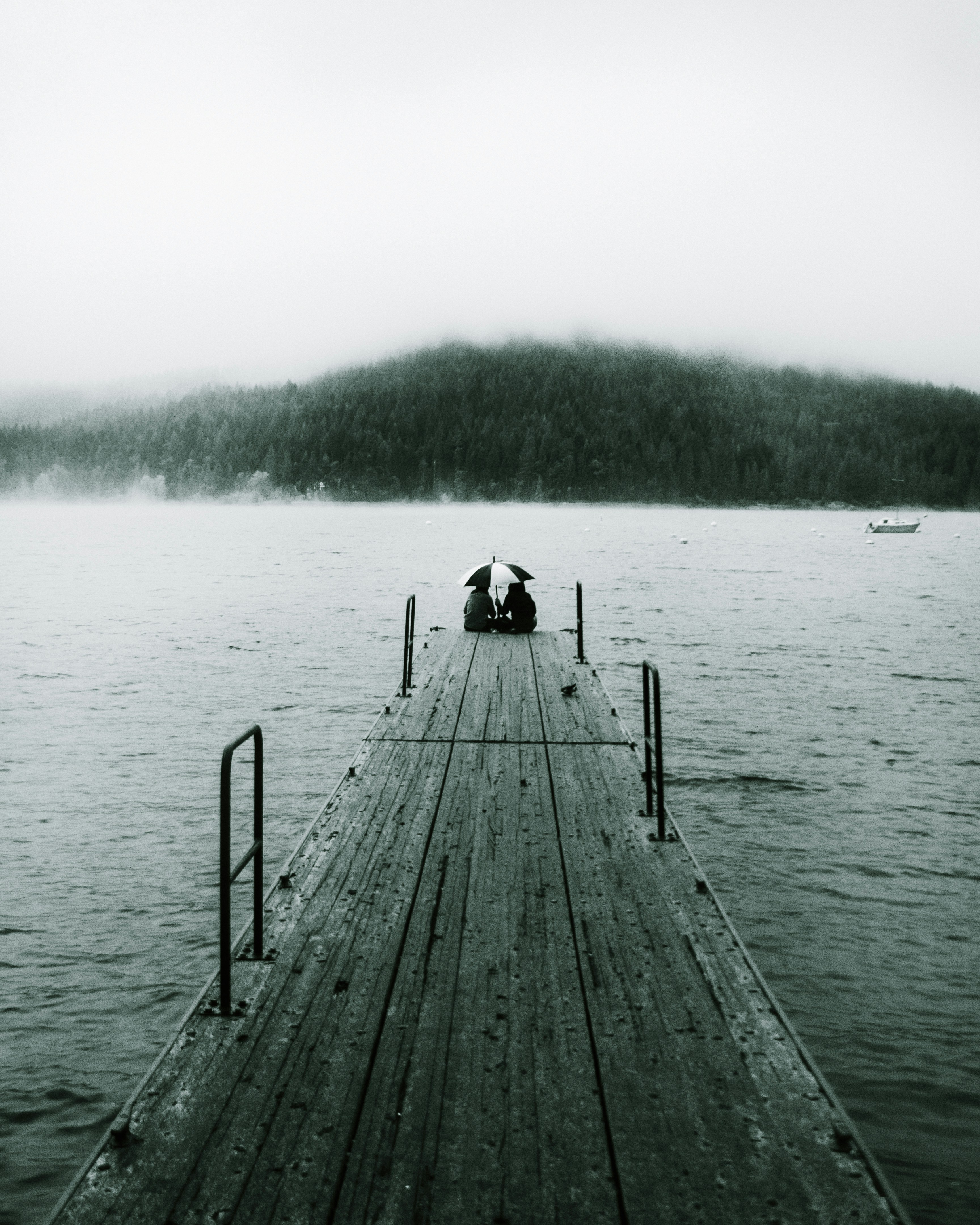 Couple seated under an umbrella on a wooden pier, surrounded by misty waters and distant hills. The scene evokes a sense of intimacy and tranquility.