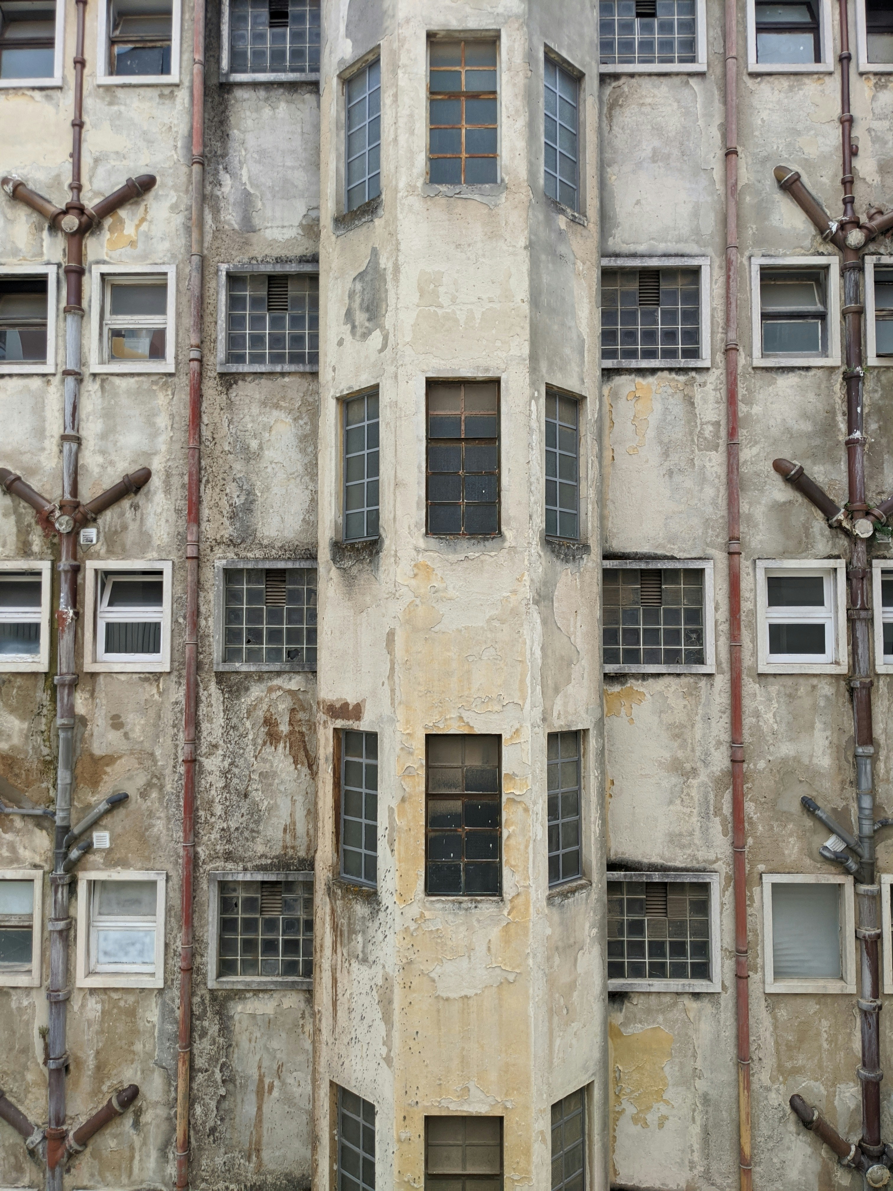 Weathered facade of a building showcasing a cylindrical structure surrounded by rectangular windows and pipes, highlighting urban decay.