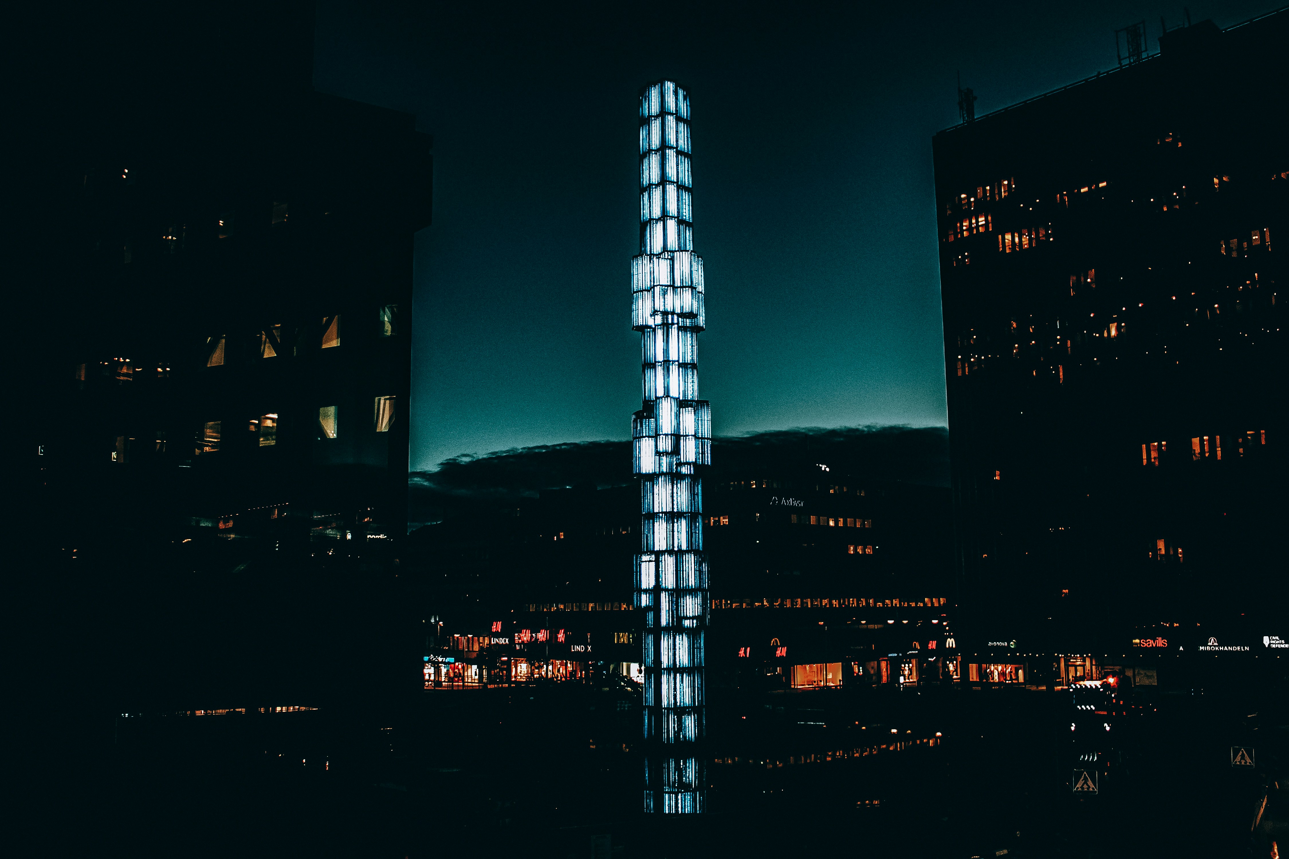 Tall, illuminated tower standing between dark skyscrapers at night.