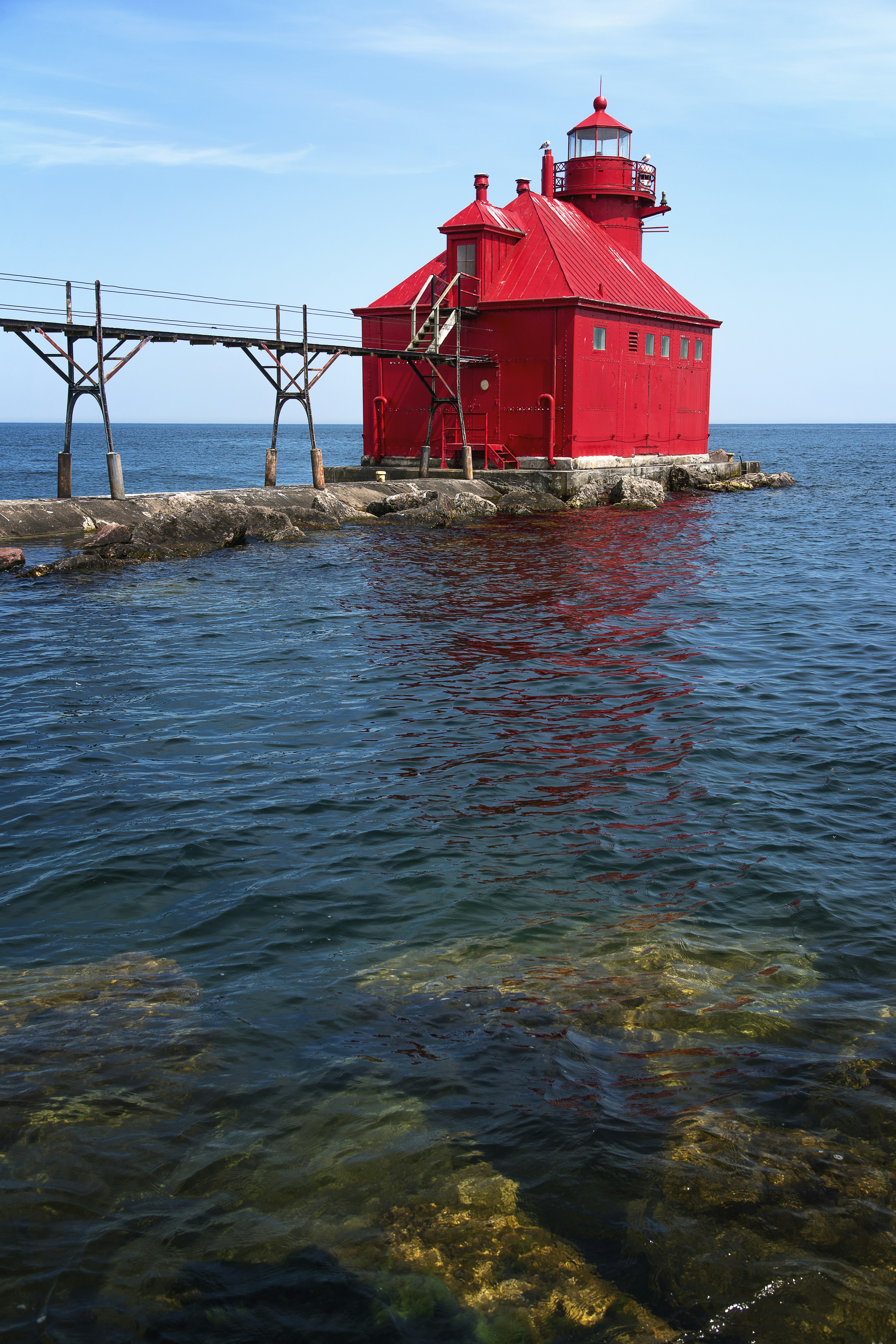 A vibrant red lighthouse stands on a rocky outcrop, connected by a wooden walkway, reflecting in the calm blue waters beneath a clear sky.
