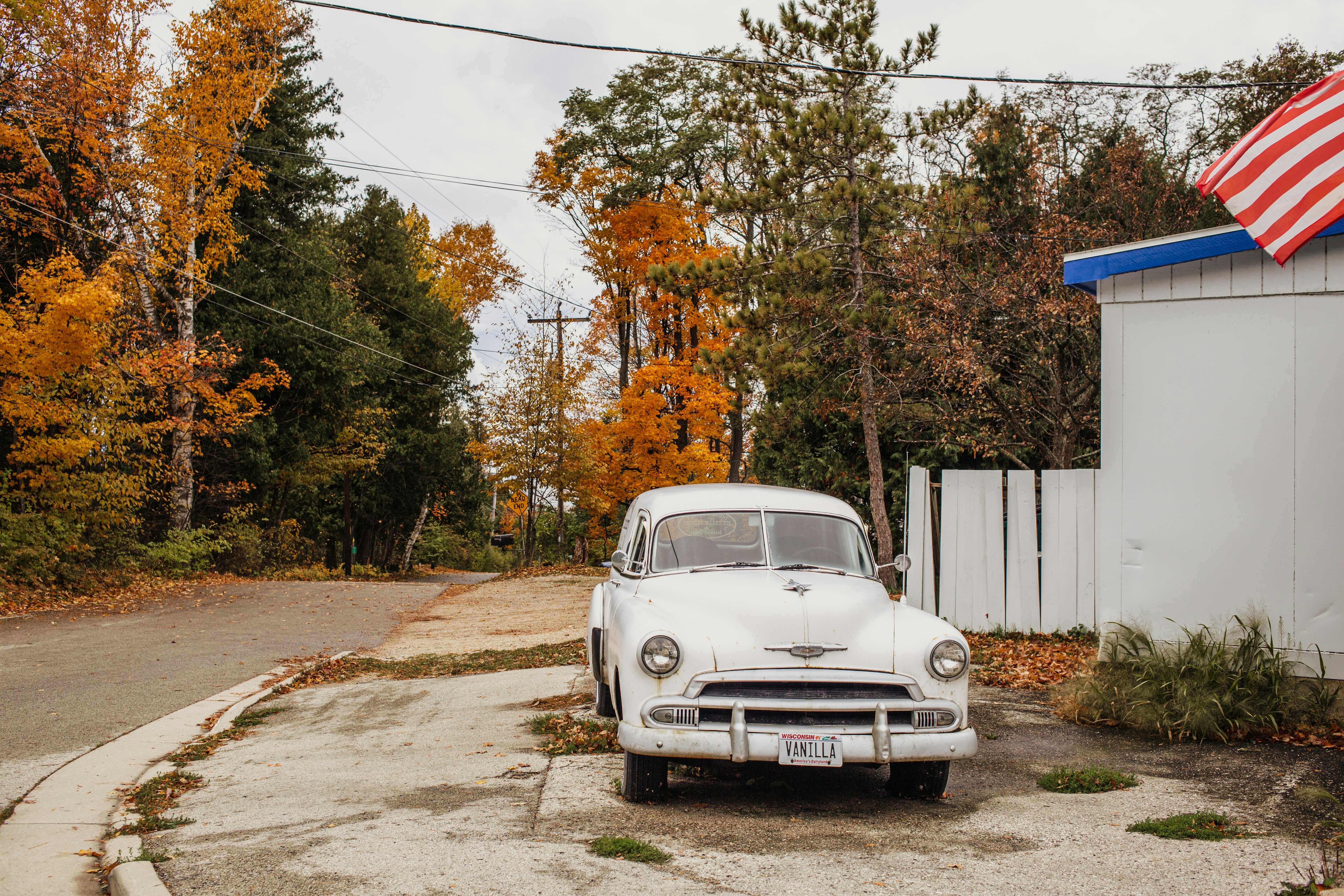 Classic white car parked beside a tree-lined road adorned with vibrant autumn foliage. An American flag flutters nearby.