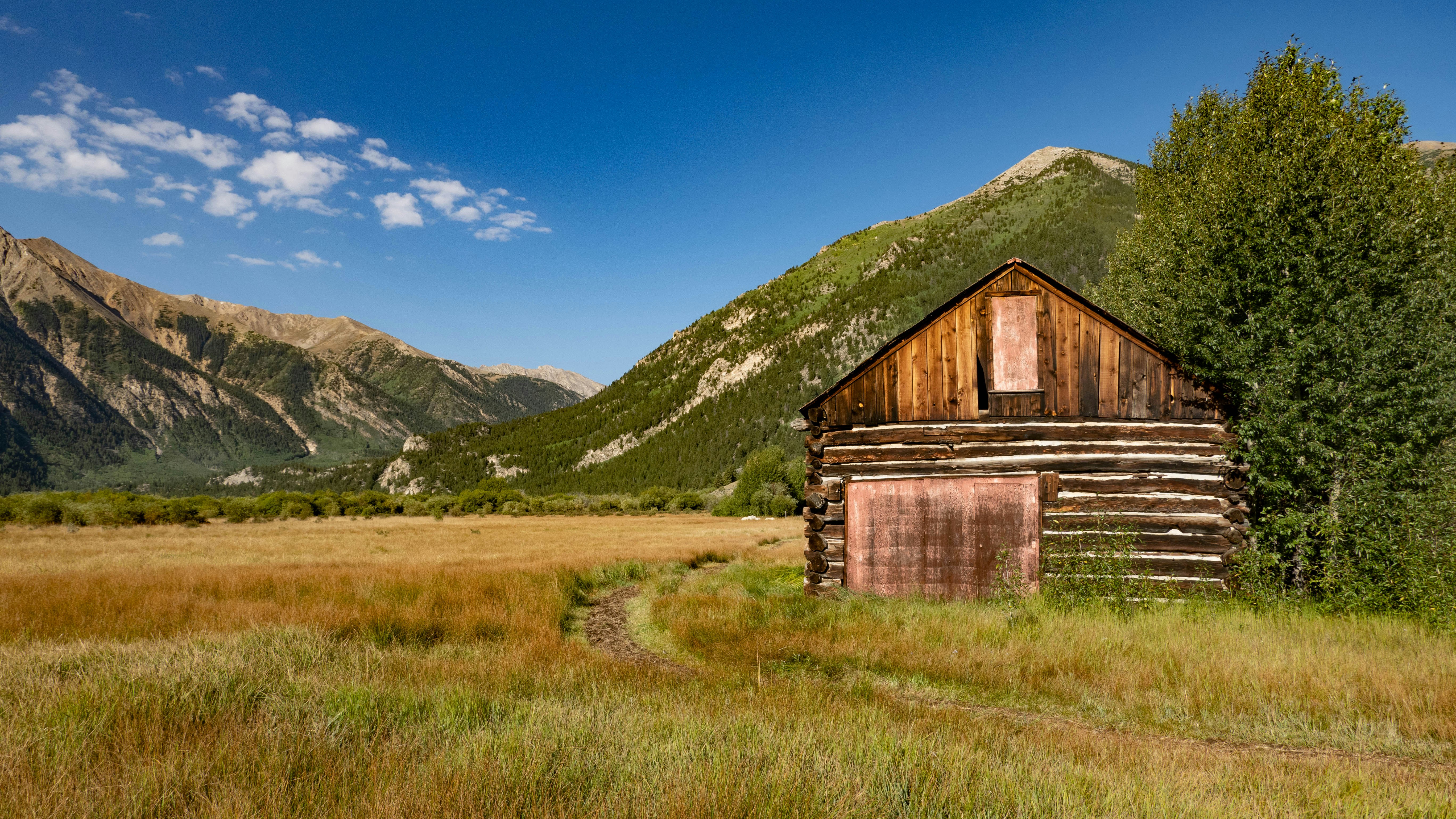 Weathered wooden barn set against a backdrop of grassy fields and majestic mountains under a clear blue sky.