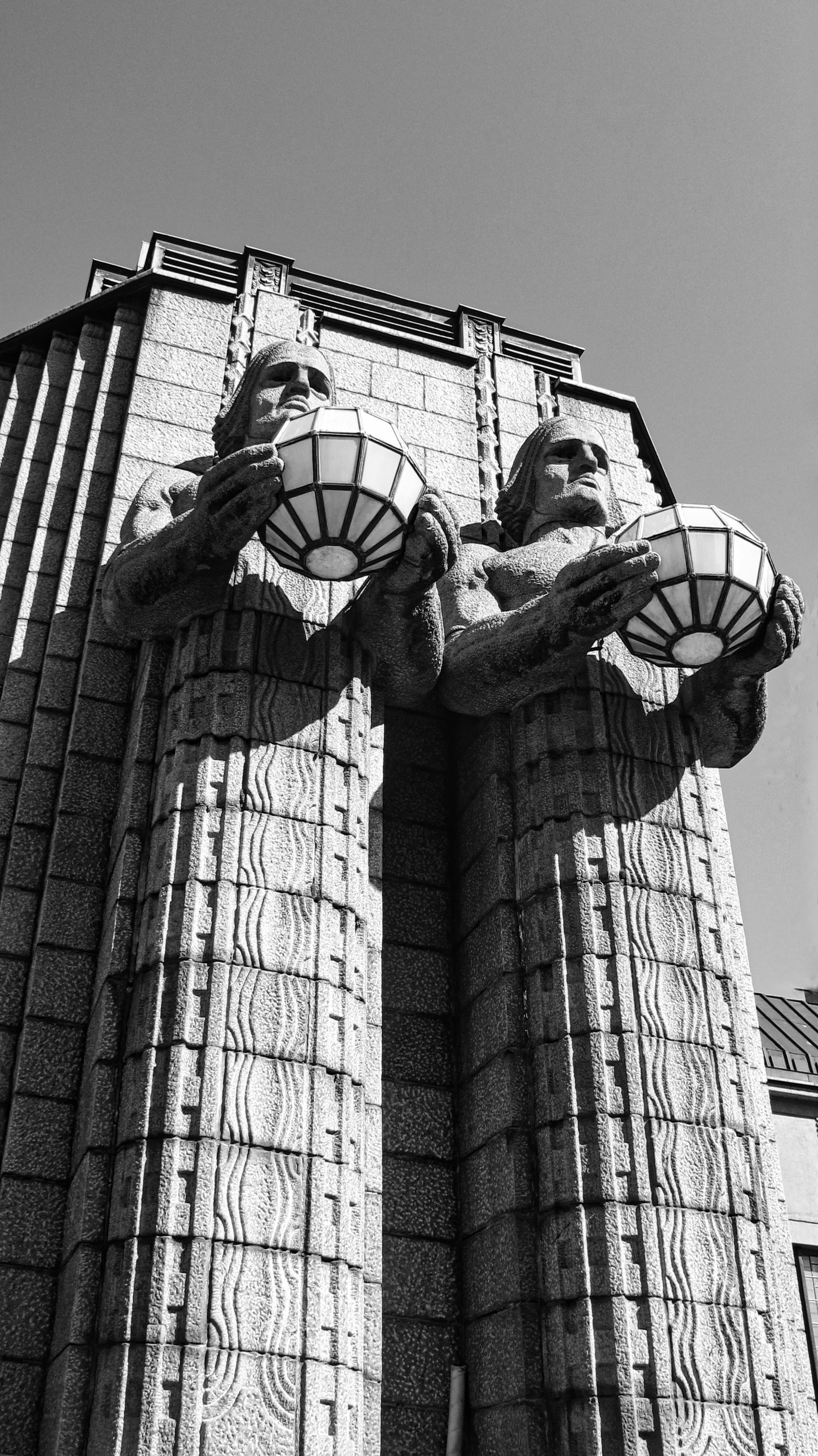 Twin stone statues grip glass lanterns atop a granite facade, photographed in stark black-and-white. The composition highlights monumental scale and intricate sculptural relief.