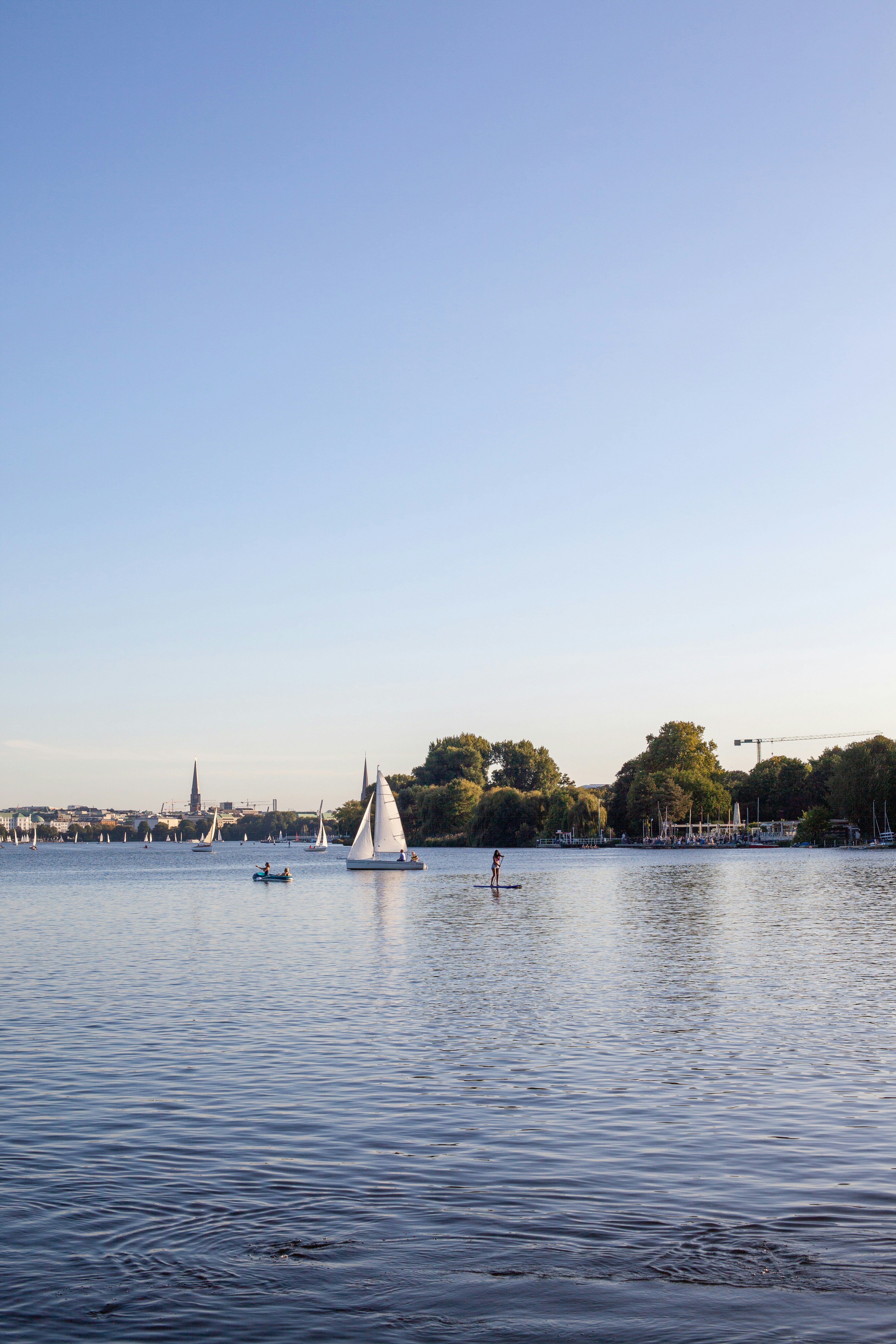 Sailboats and paddleboarders enjoying a tranquil lake under a clear blue sky. The scene captures the essence of outdoor leisure activities.
