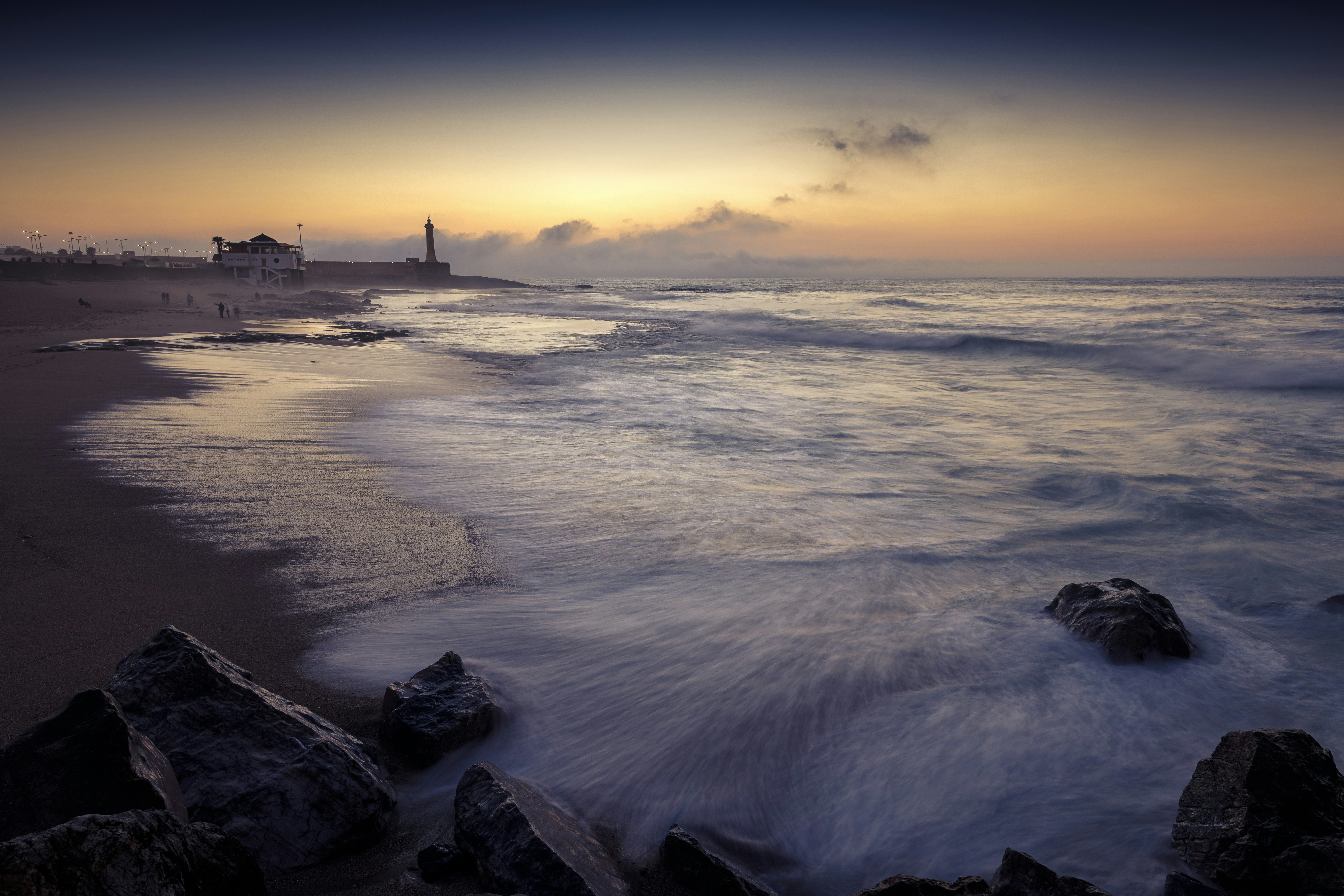 Gentle waves lap against the sandy beach at twilight, with a lighthouse standing sentinel in the distance. The soft hues of sunset blend seamlessly with the horizon.