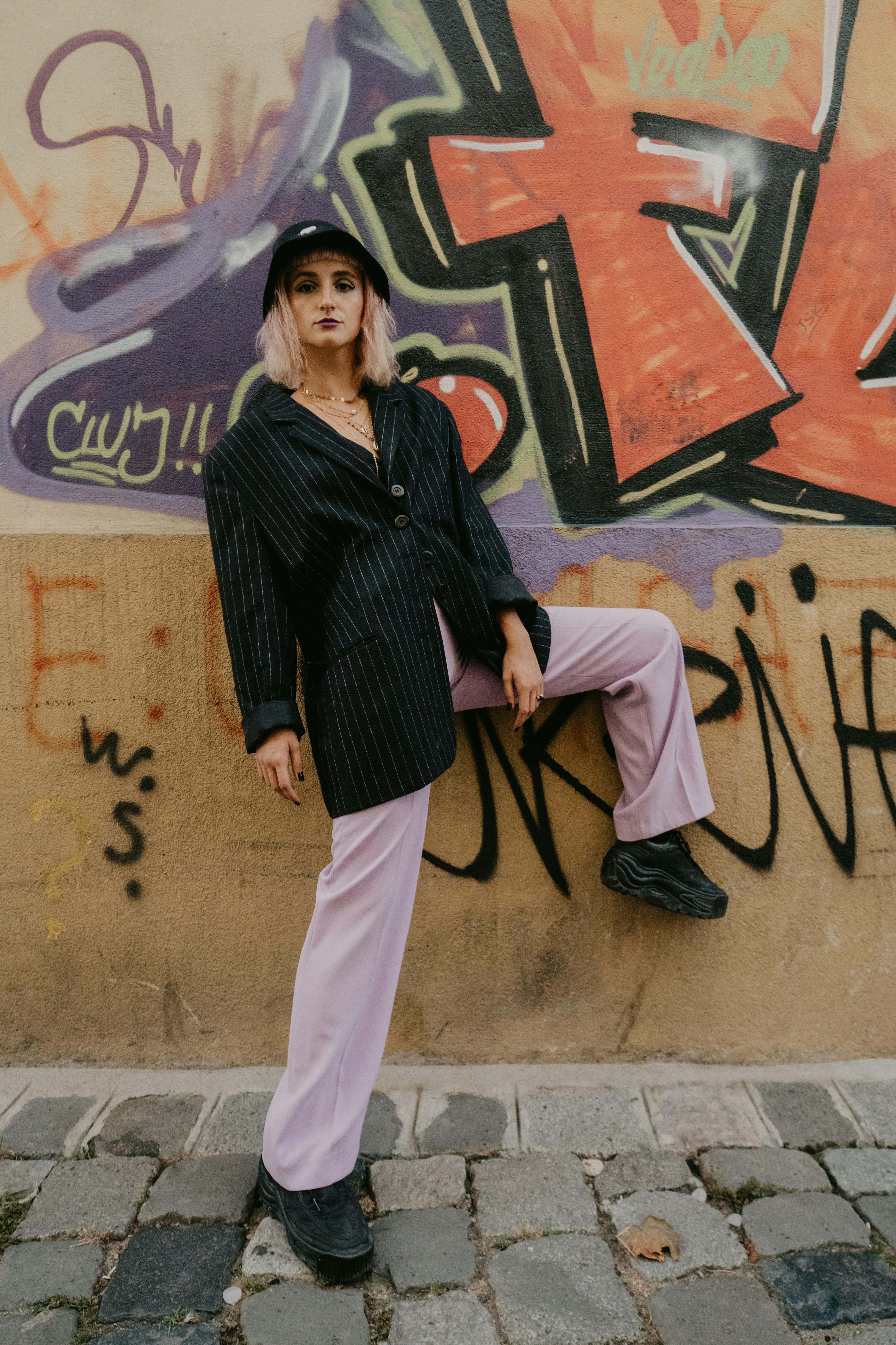 Fashion-forward individual posing against a vibrant graffiti backdrop, showcasing a unique blend of styles with a pinstripe blazer and pastel trousers.