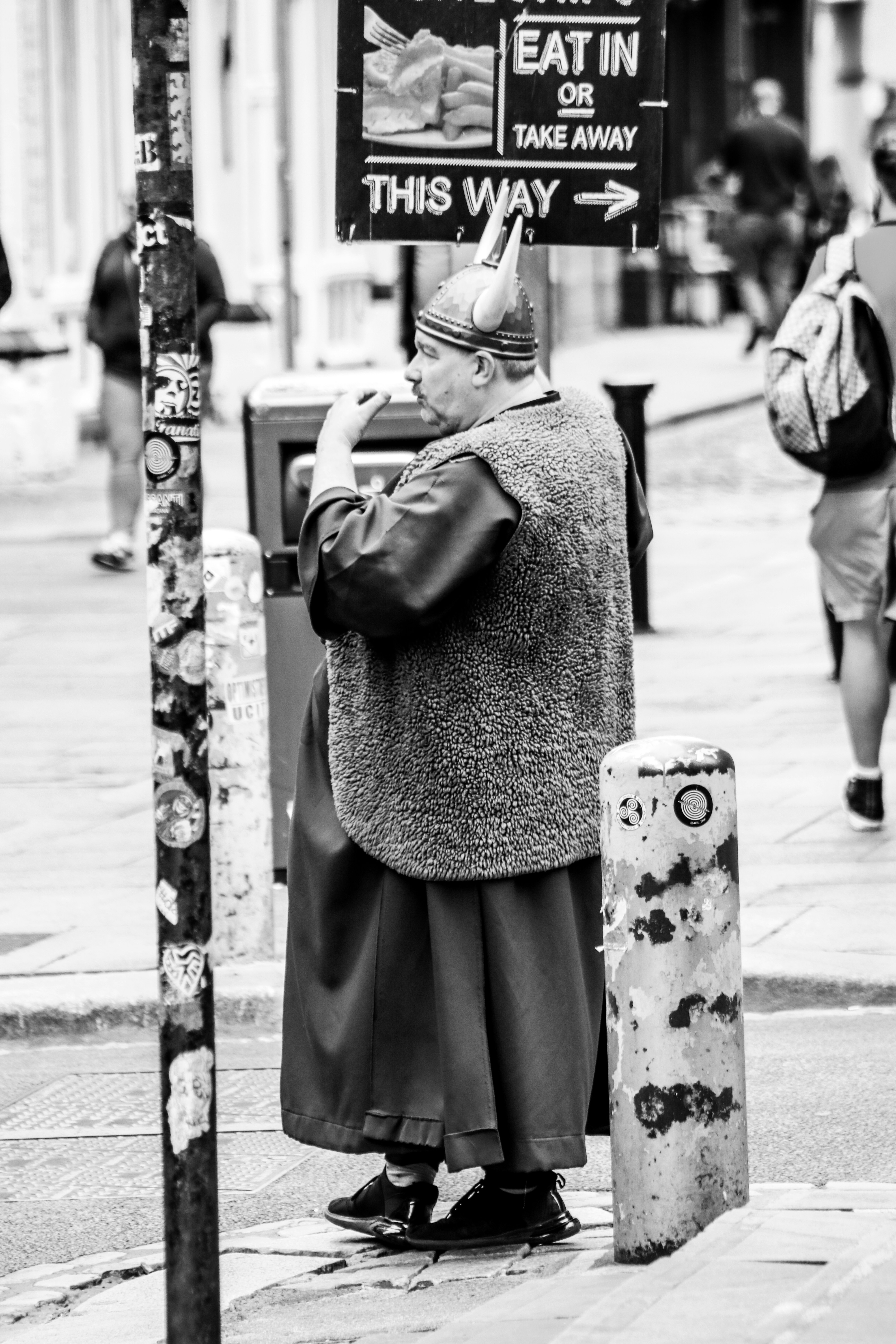 A man in a unique outfit stands near a street sign, smoking thoughtfully amidst a bustling city scene.