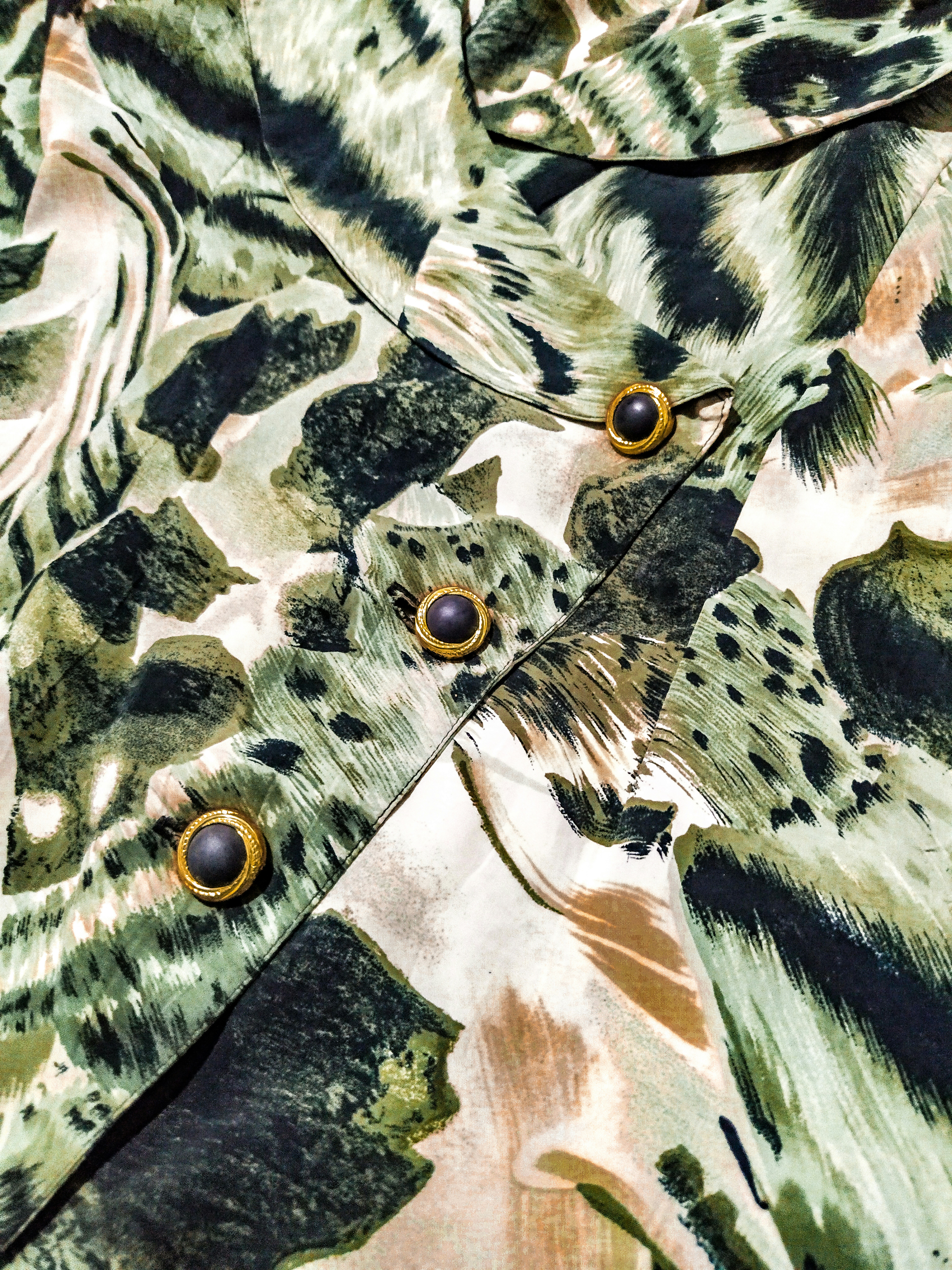 Close-up of a patterned fabric featuring an intricate blend of greens and earth tones, accented by decorative buttons. The design showcases a fusion of natural elements.