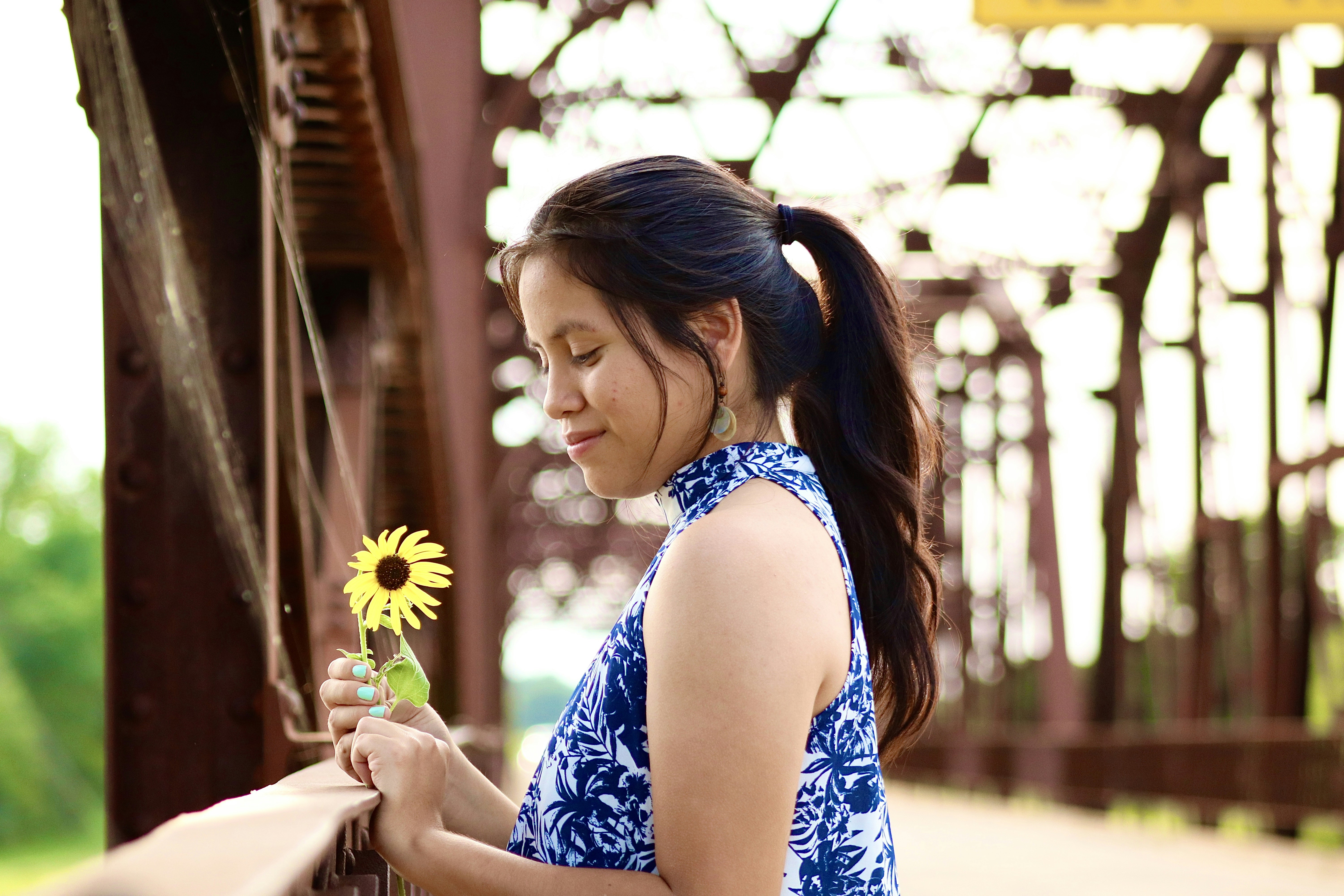 A young woman in a floral blue dress holds a sunflower while leaning on a weathered bridge railing, basking in the serene atmosphere.