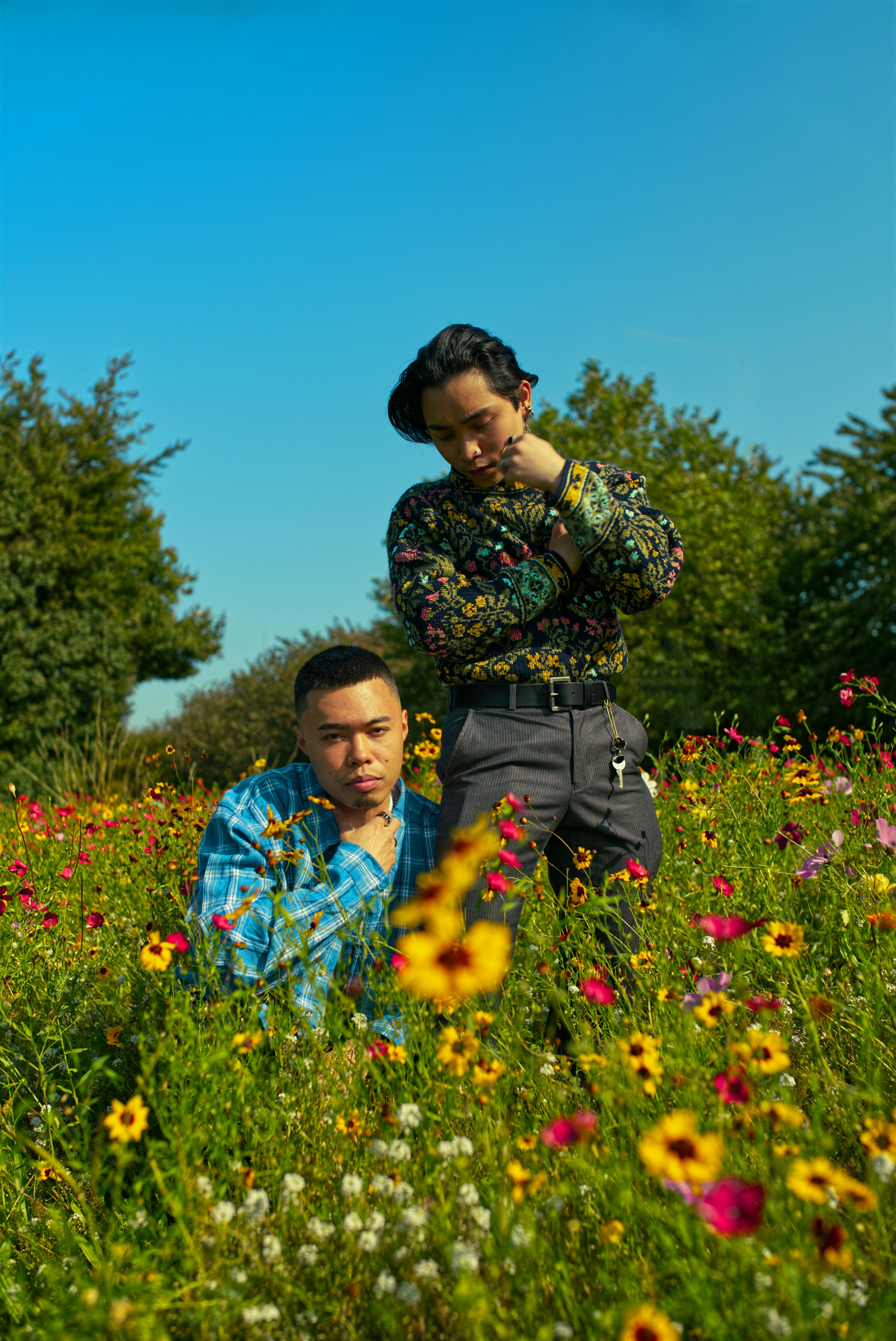 Two individuals striking poses in a colorful wildflower field, showcasing unique fashion choices and expressions. Bright flowers surround them, enhancing the lively atmosphere.