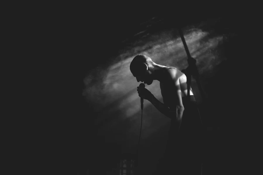 A stark black-and-white scene shows a shirtless singer bent over a microphone in dramatic side light. The surrounding stage falls into deep shadow, emphasizing the figure’s silhouette and mood.