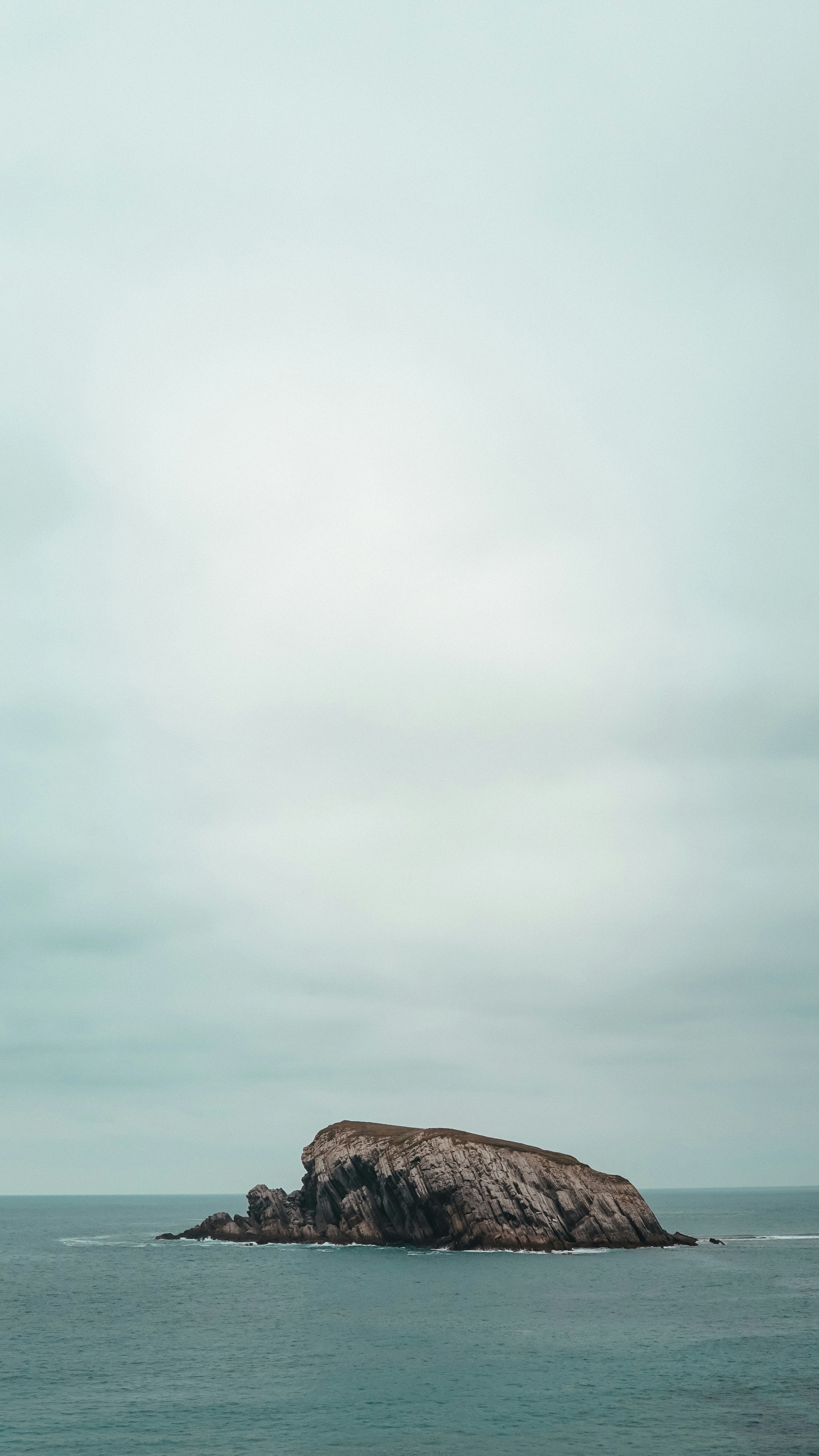 A solitary rock formation rises from the calm ocean, surrounded by soft, overcast skies. The scene evokes a sense of peace and solitude.