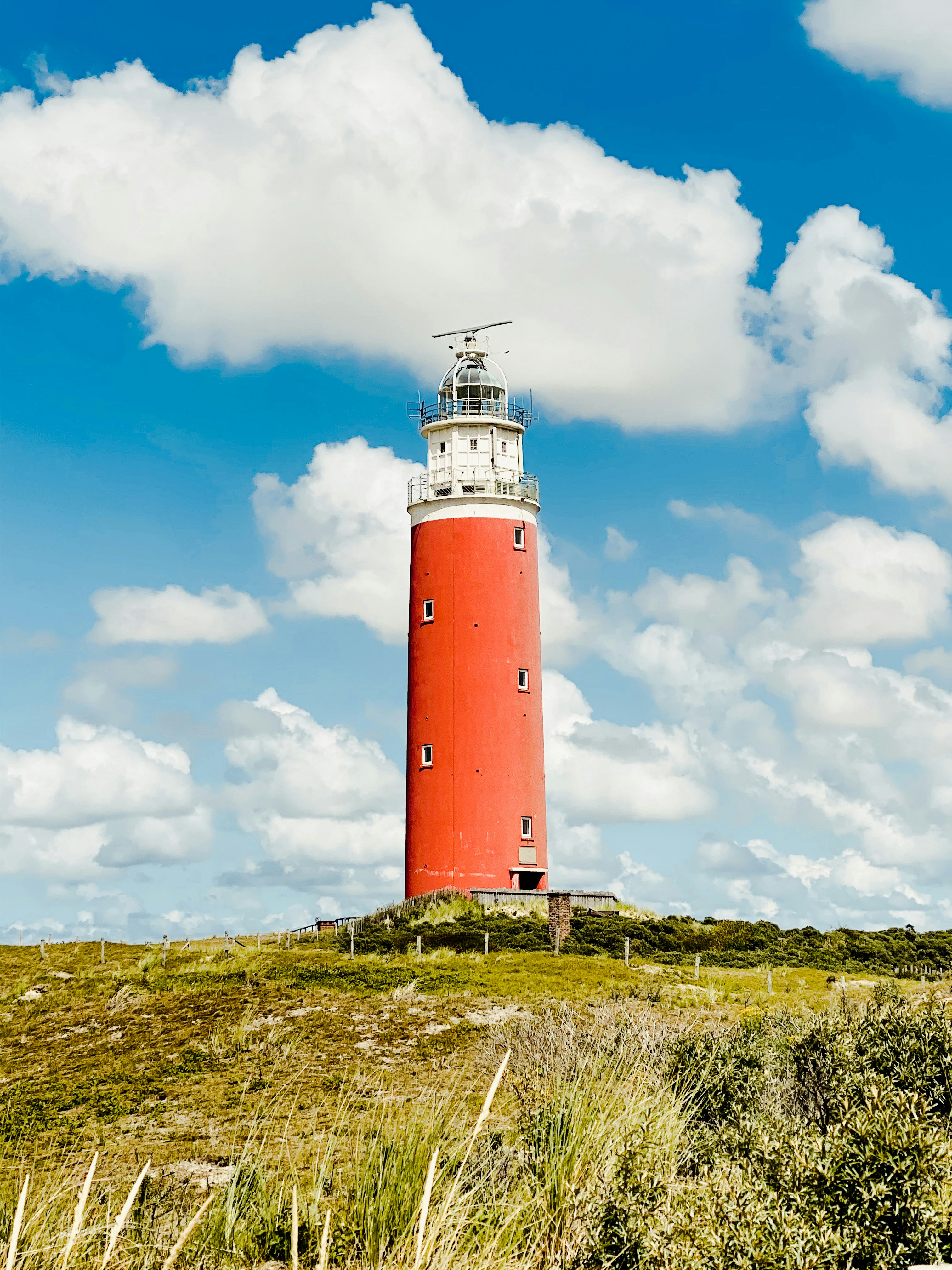 A striking red lighthouse stands tall against a backdrop of fluffy white clouds and a vibrant blue sky, surrounded by lush greenery.