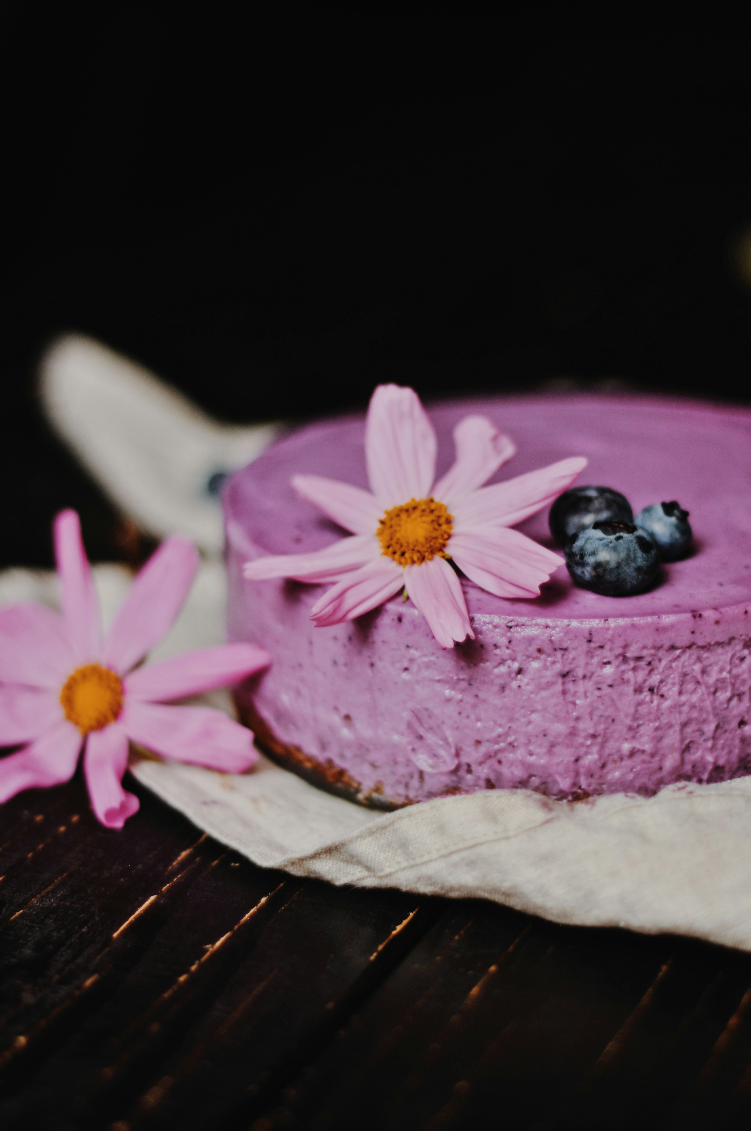 A vibrant purple cheesecake garnished with daisies and blueberries, set against a dark wooden background. The arrangement emphasizes the dessert's freshness and appeal.